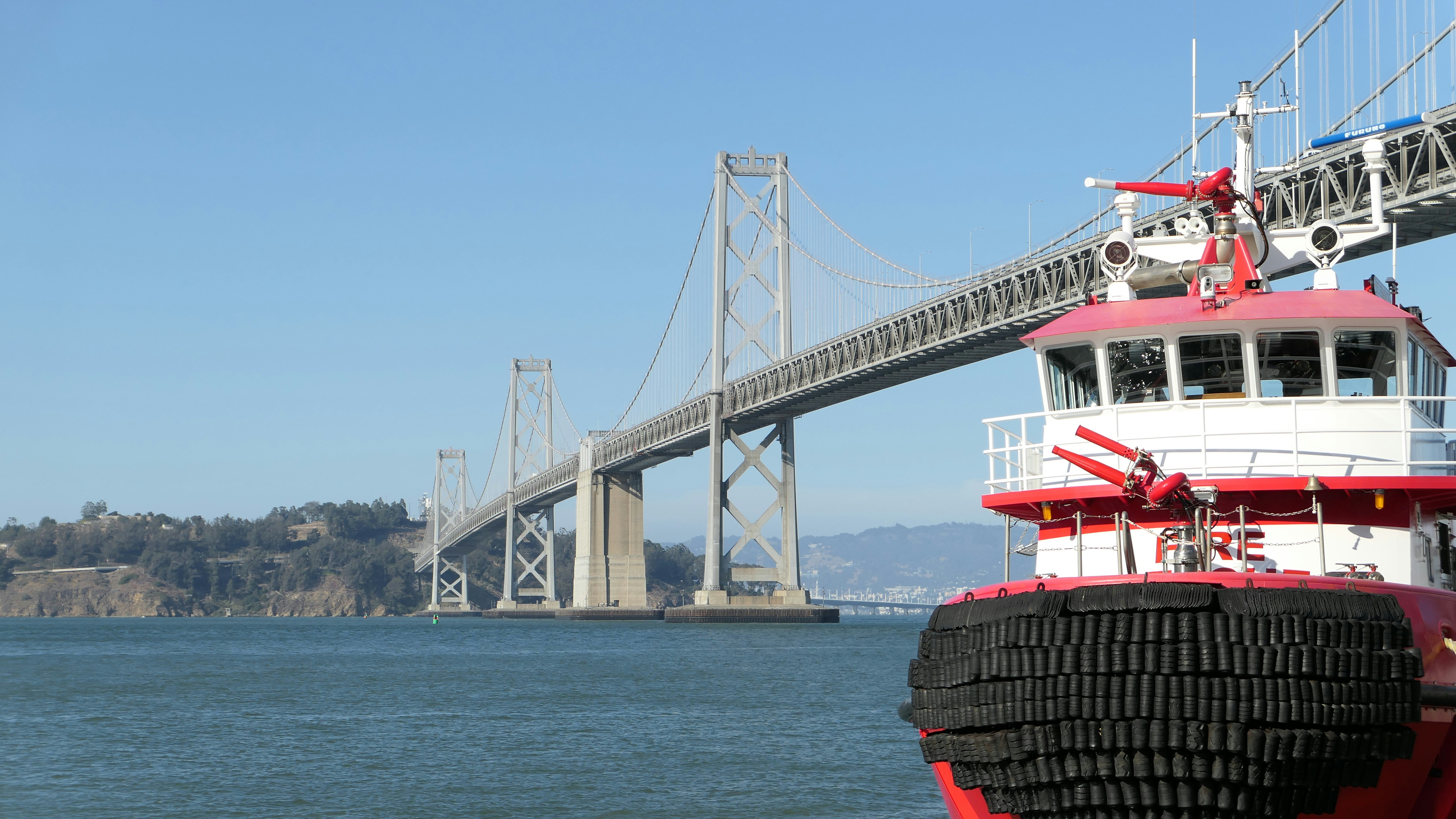 Photo shows a red and white tugboat with large black tires in the foreground as the Bay Bridge extends across calm water beneath a clear blue sky.
