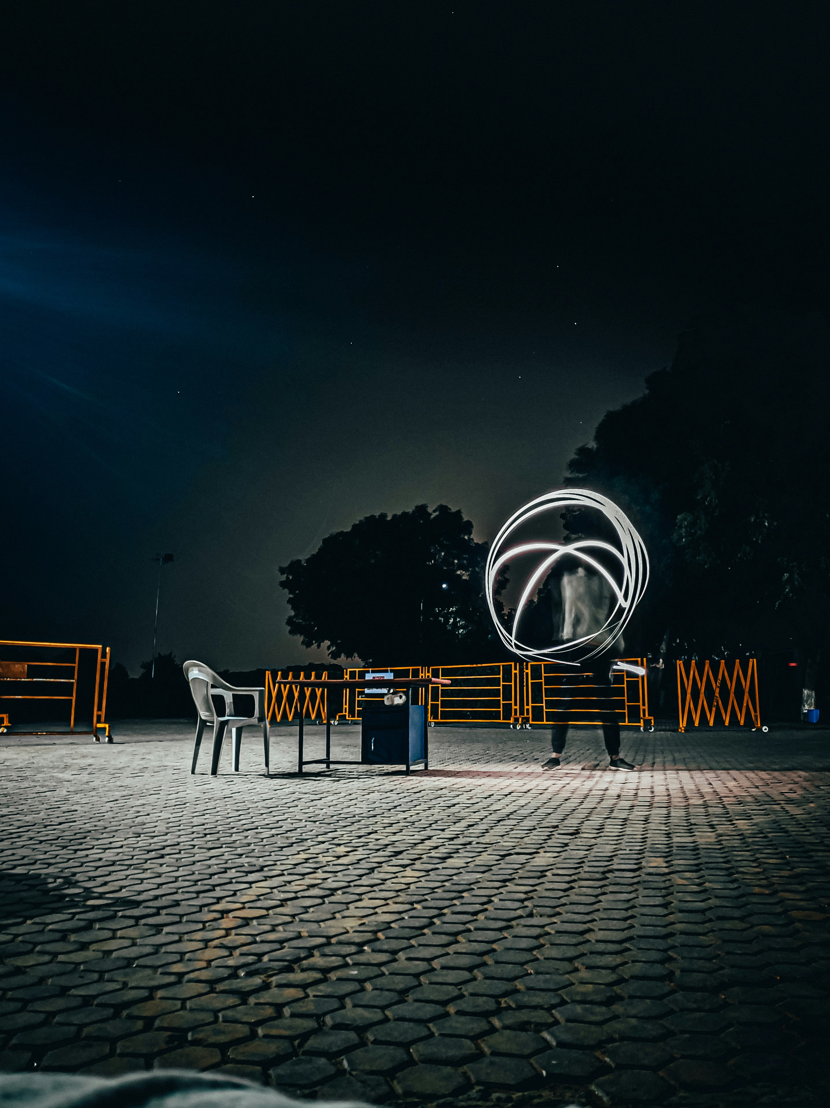 A person creates light trails with a glowing orb in a nighttime setting, surrounded by a chair and barriers. The scene captures the essence of movement and creativity.