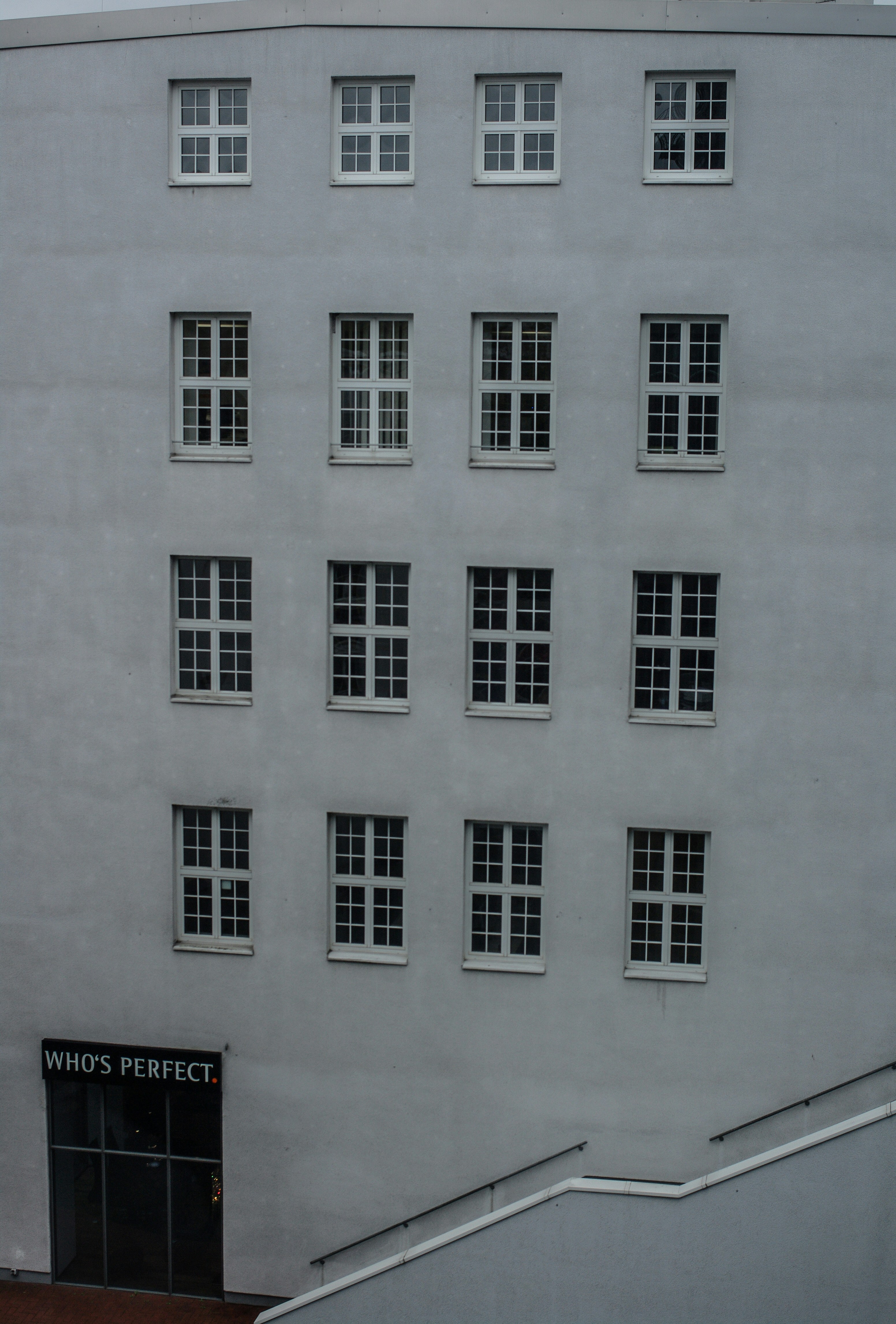 Gray facade with a precise grid of rectangular windows dominates the frame. A doorway at bottom left bears the text 'WHO'S PERFECT,' with a metal railing looping up the steps.