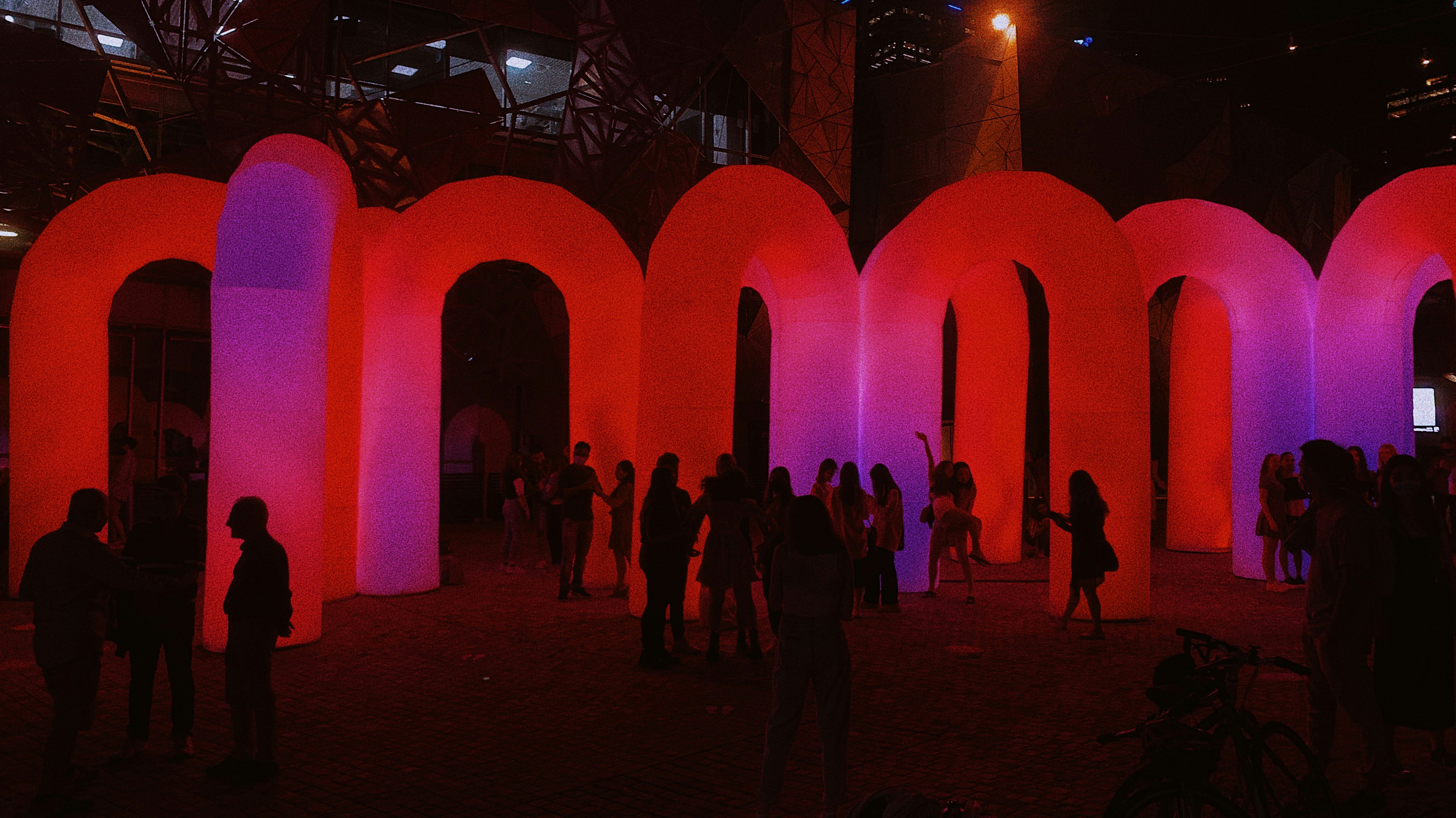 Silhouettes of people against vibrant red and pink illuminated arches at night.