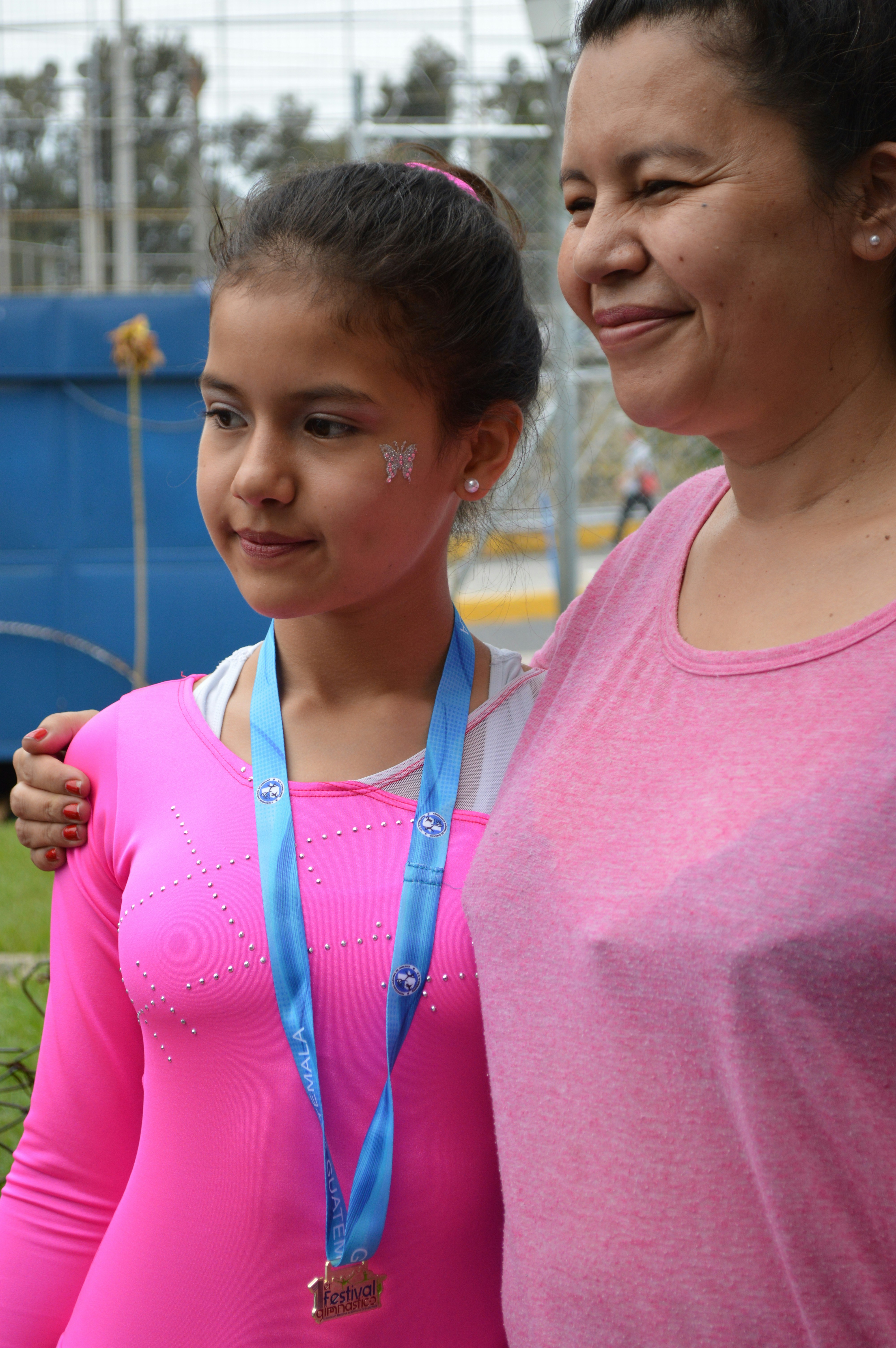 A young gymnast in a vibrant pink outfit proudly displays her medal alongside her supportive mother, set against a backdrop of a sports venue.