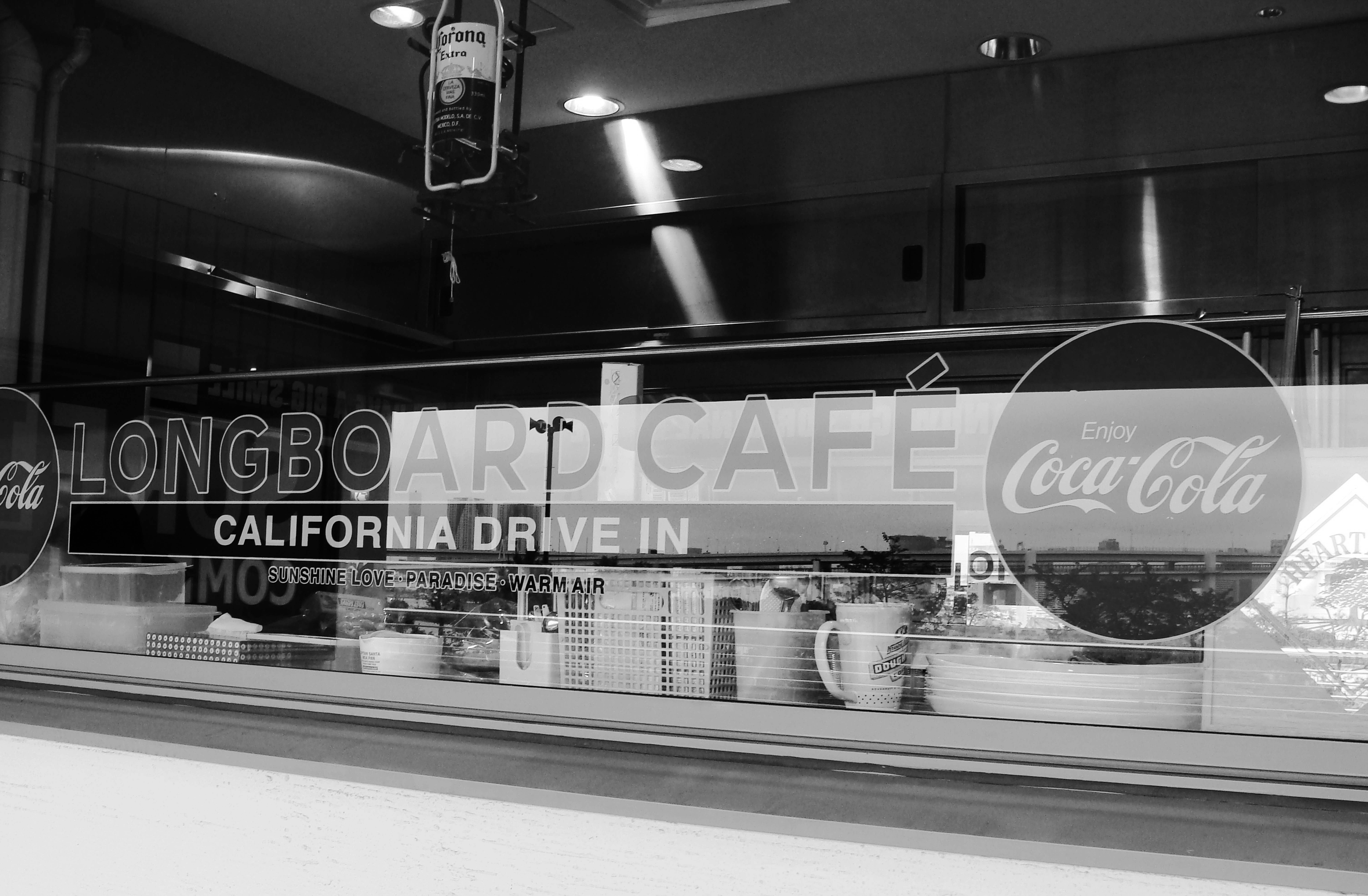 Black and white illustration of a café window featuring the name 'LONGBOARD CAFÉ' alongside a Coca-Cola advertisement. The scene evokes a nostalgic drive-in atmosphere.