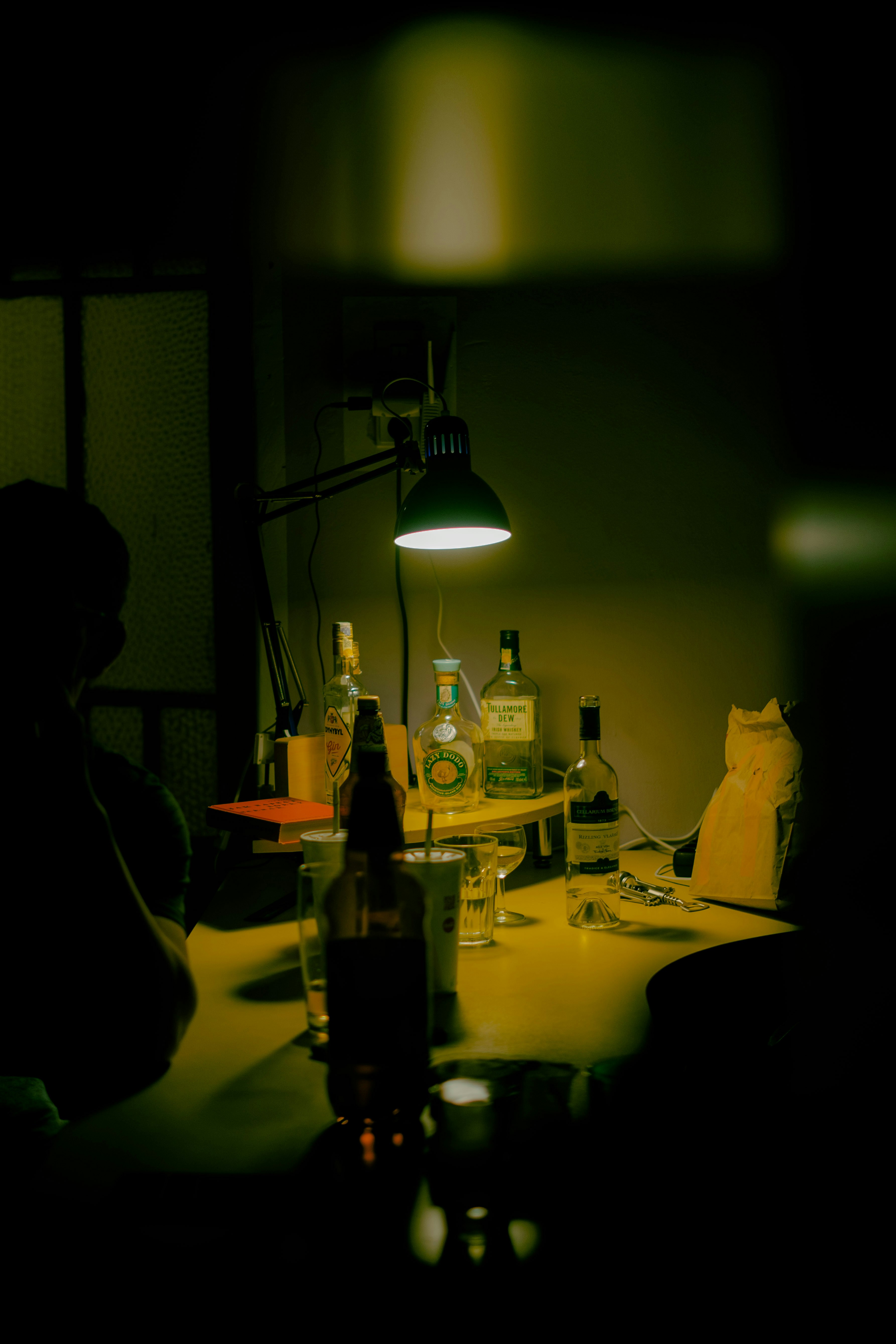 A dimly lit table adorned with various bottles and glasses, illuminated by a green desk lamp, creating an intimate atmosphere.