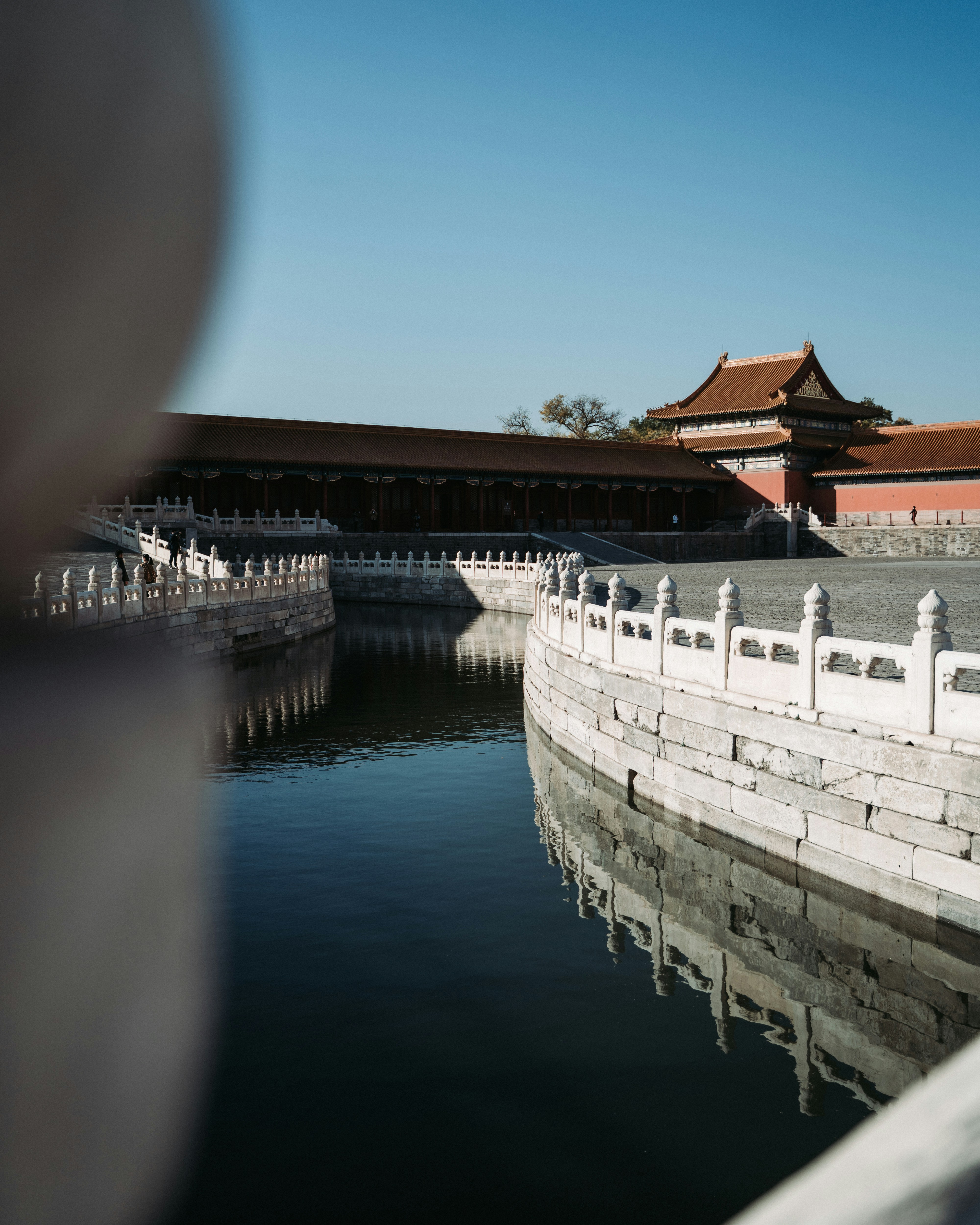 Curved stone railing frames a tranquil waterway leading to traditional Asian architecture under a clear blue sky.