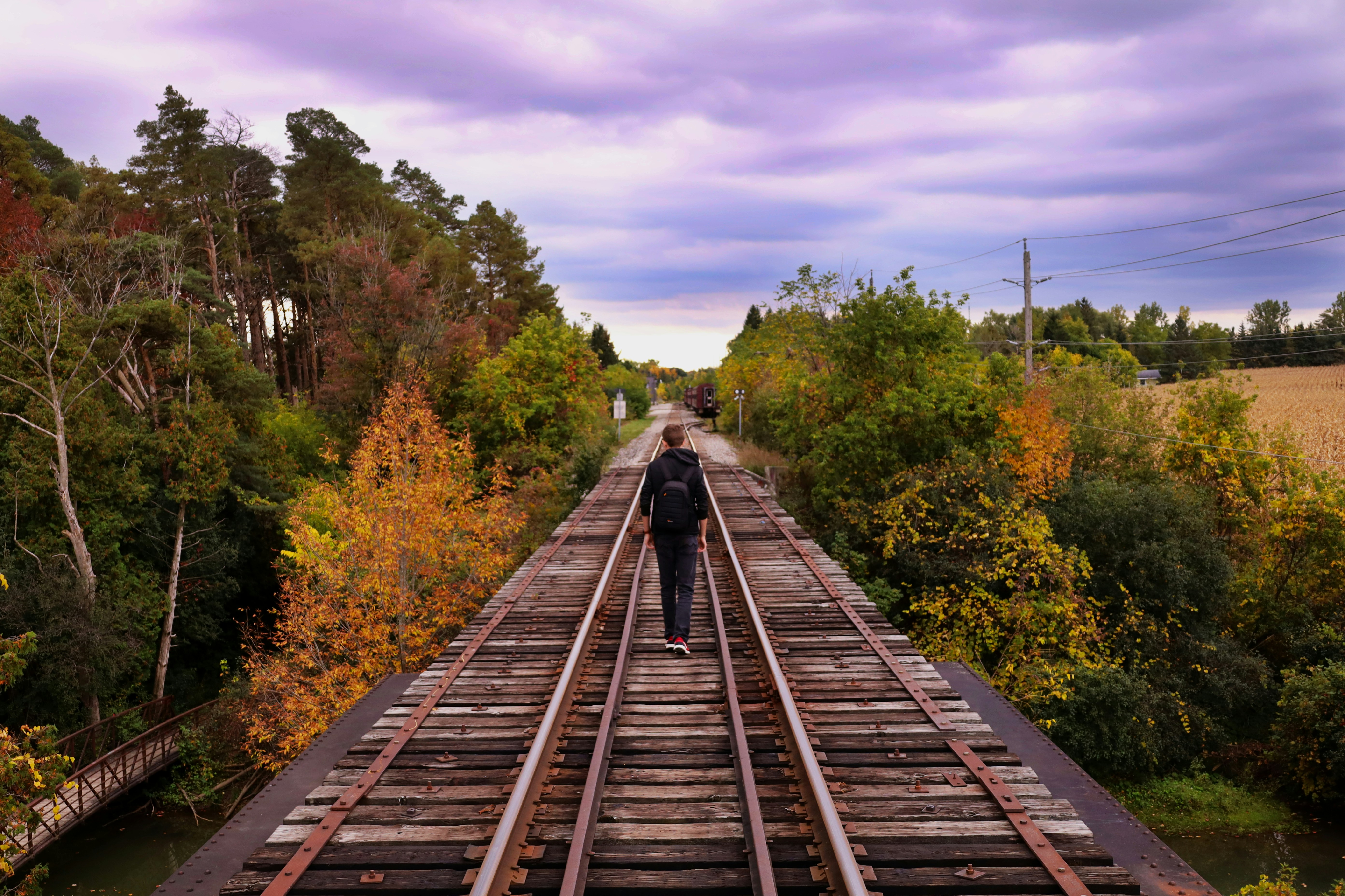 A solitary figure walks along an abandoned railway bridge, surrounded by vibrant autumn foliage and a moody sky.