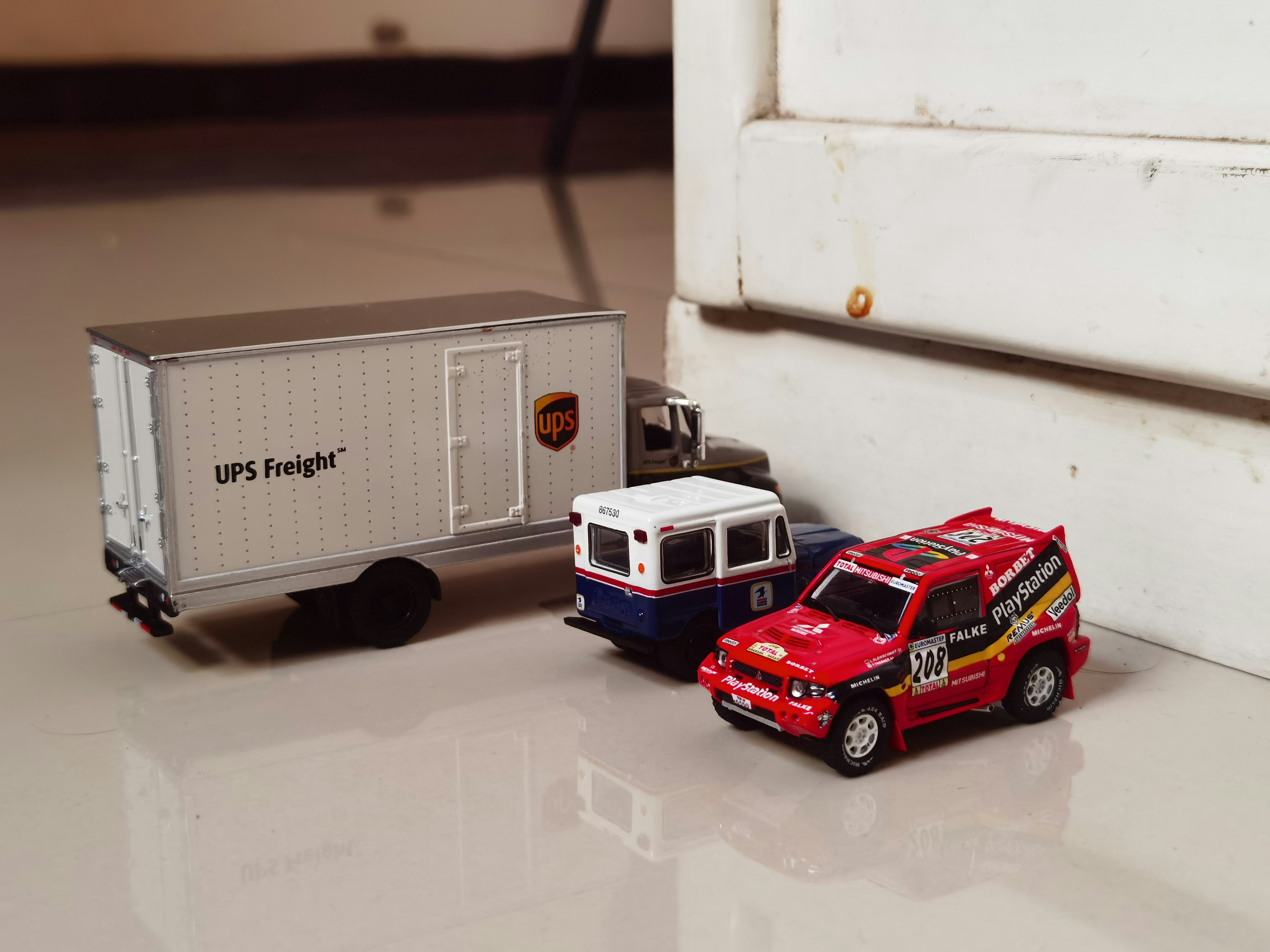 Toy vehicles—UPS Freight trailer, blue-white cab, and a red rally car—pose on a glossy tiled floor beside a white cabinet.