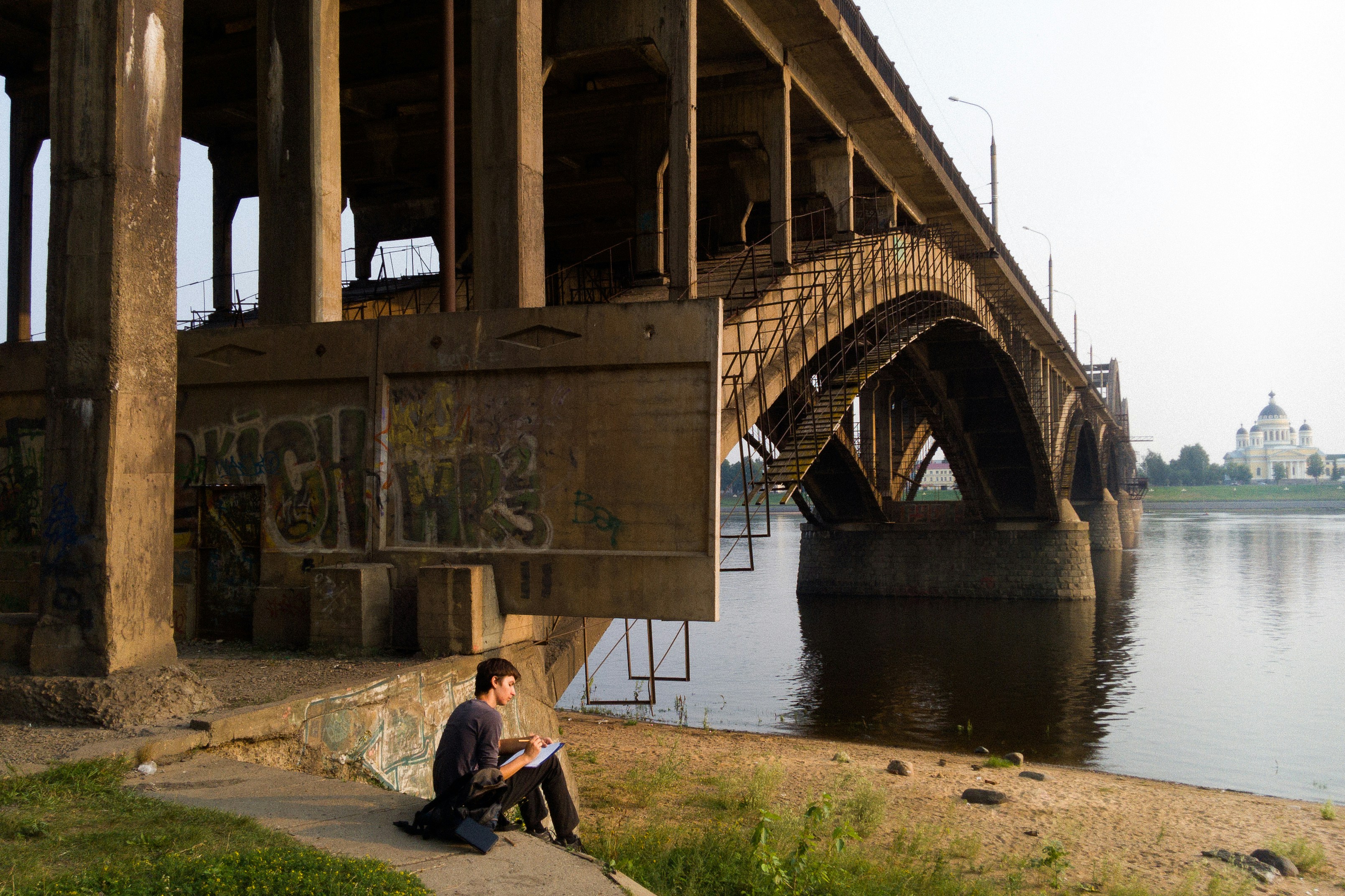 Person sitting on a riverbank beneath a large, arched bridge with a distant view of a building across the water.