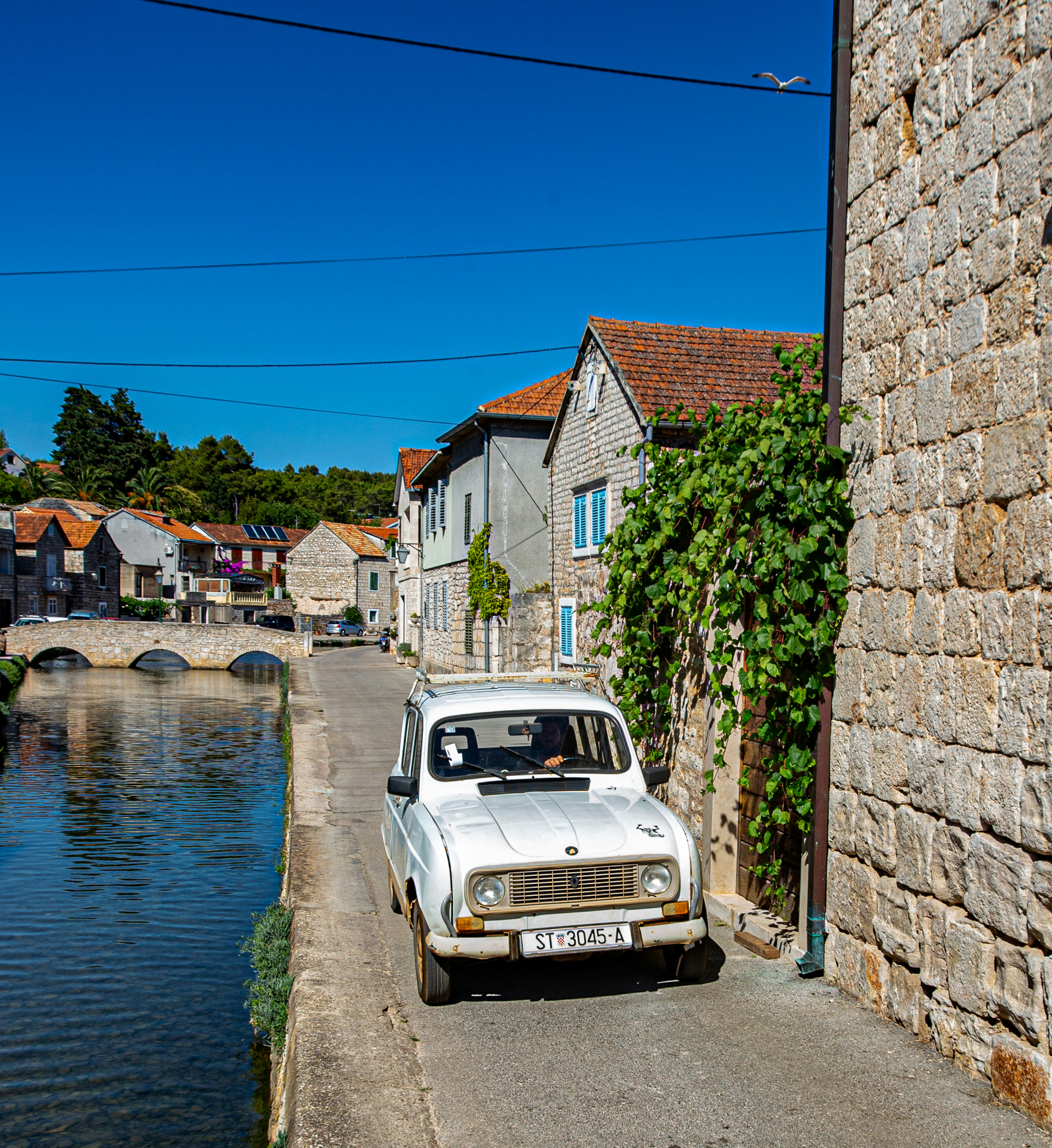 Riverside Charm: A Vintage White Sedan Anchors a Sunlit Village Lane