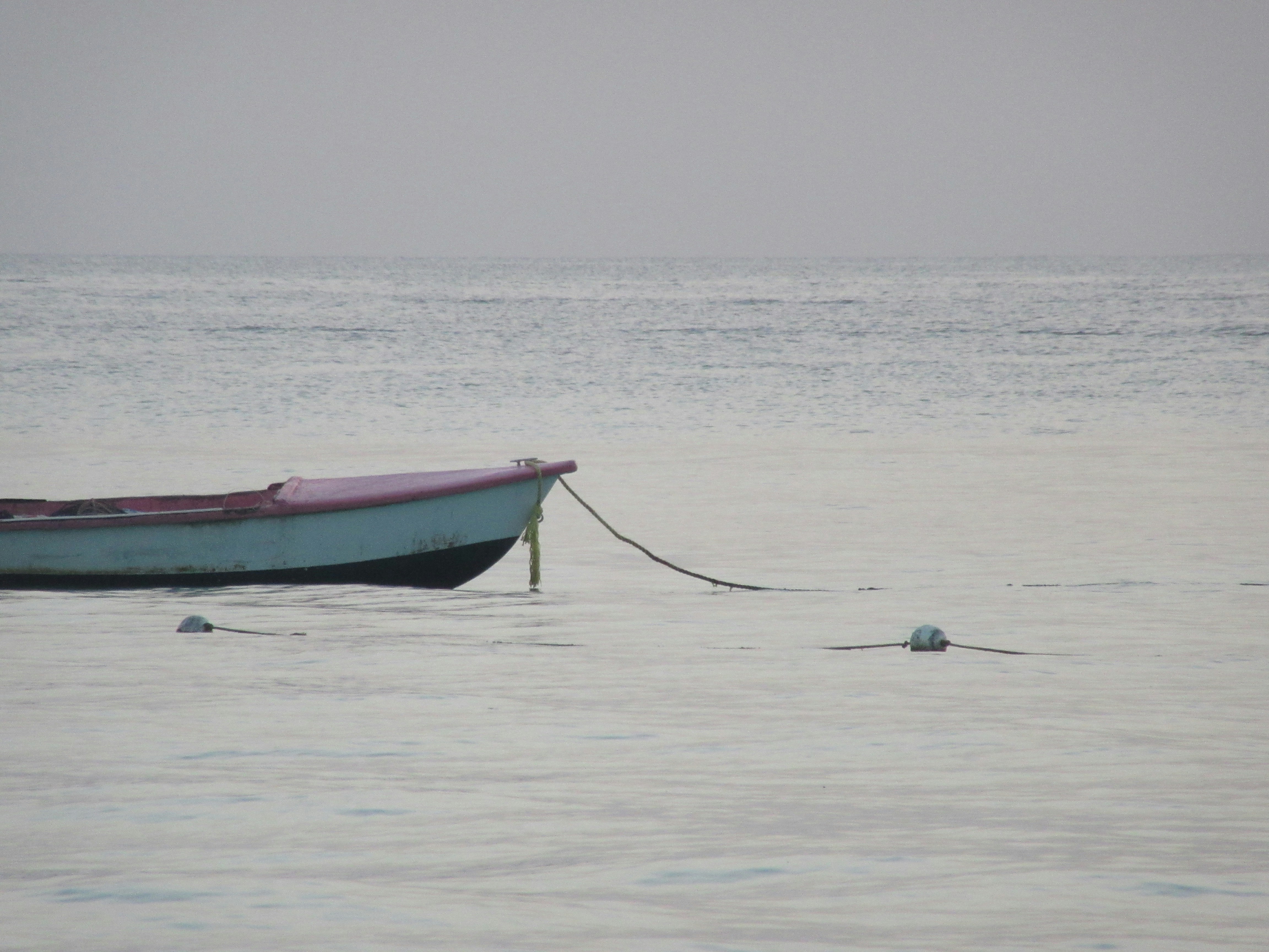 A tranquil seascape photograph of a small boat moored by a rope to a buoy in calm waters under a pale, overcast sky.