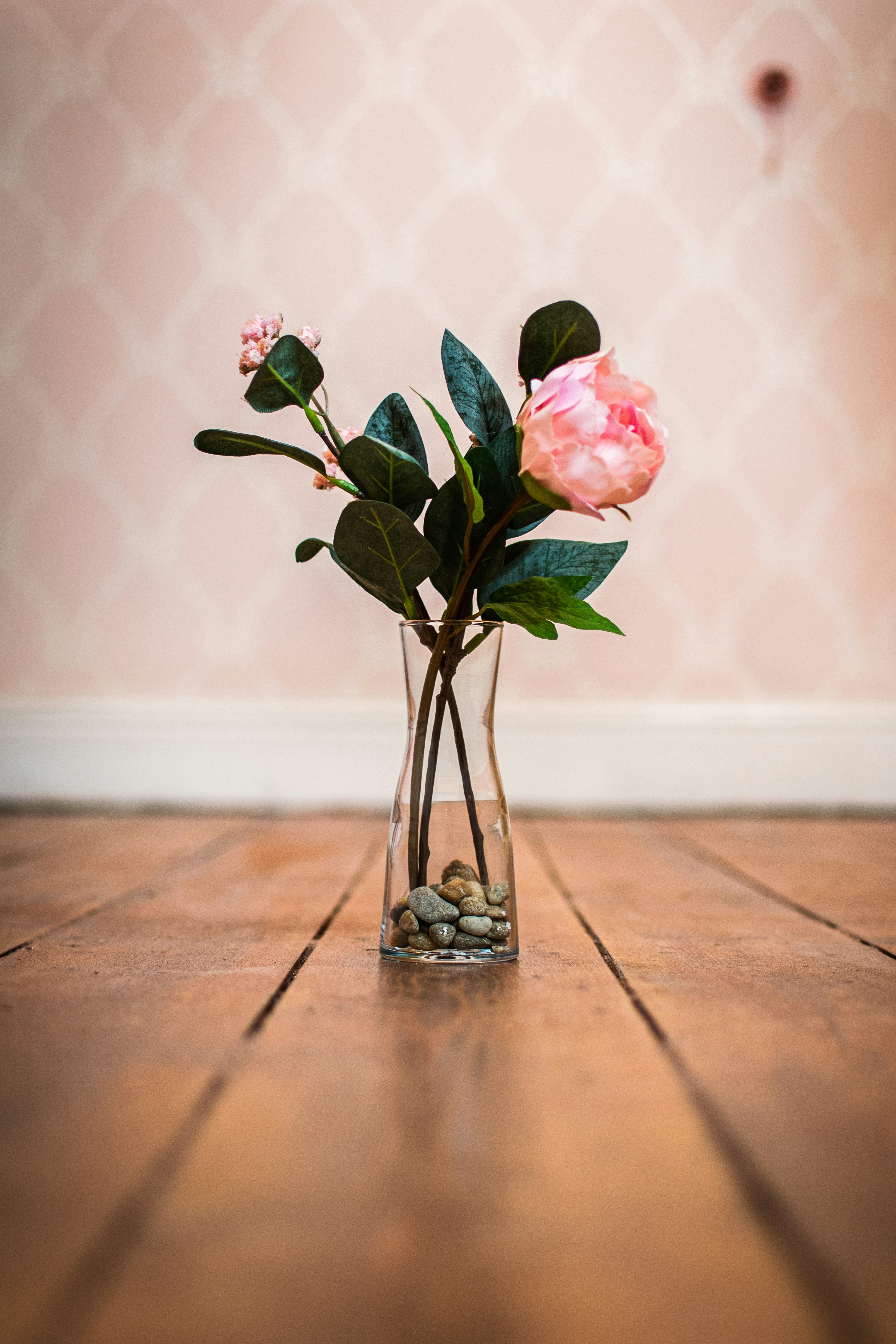A delicate pink flower arrangement in a slender glass vase, resting on a wooden floor with pebbles at the base. The soft pink wallpaper provides a gentle backdrop.