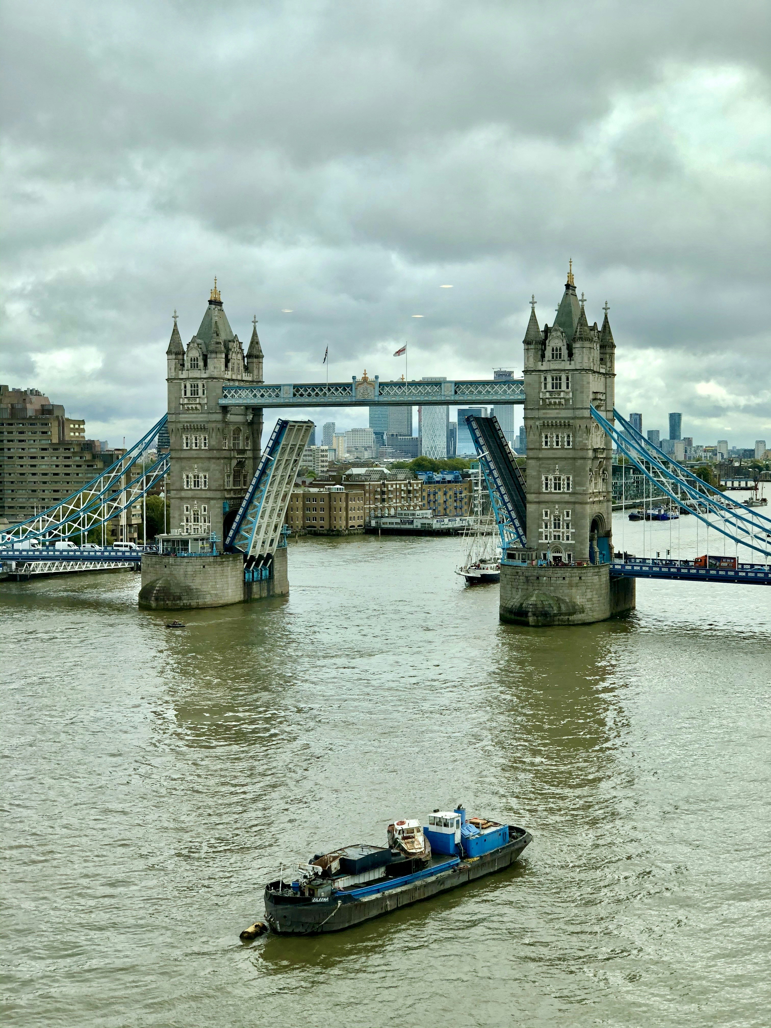 Tower Bridge spanning the River Thames, with a boat navigating beneath its iconic arches. The cloudy sky adds a dramatic backdrop.
