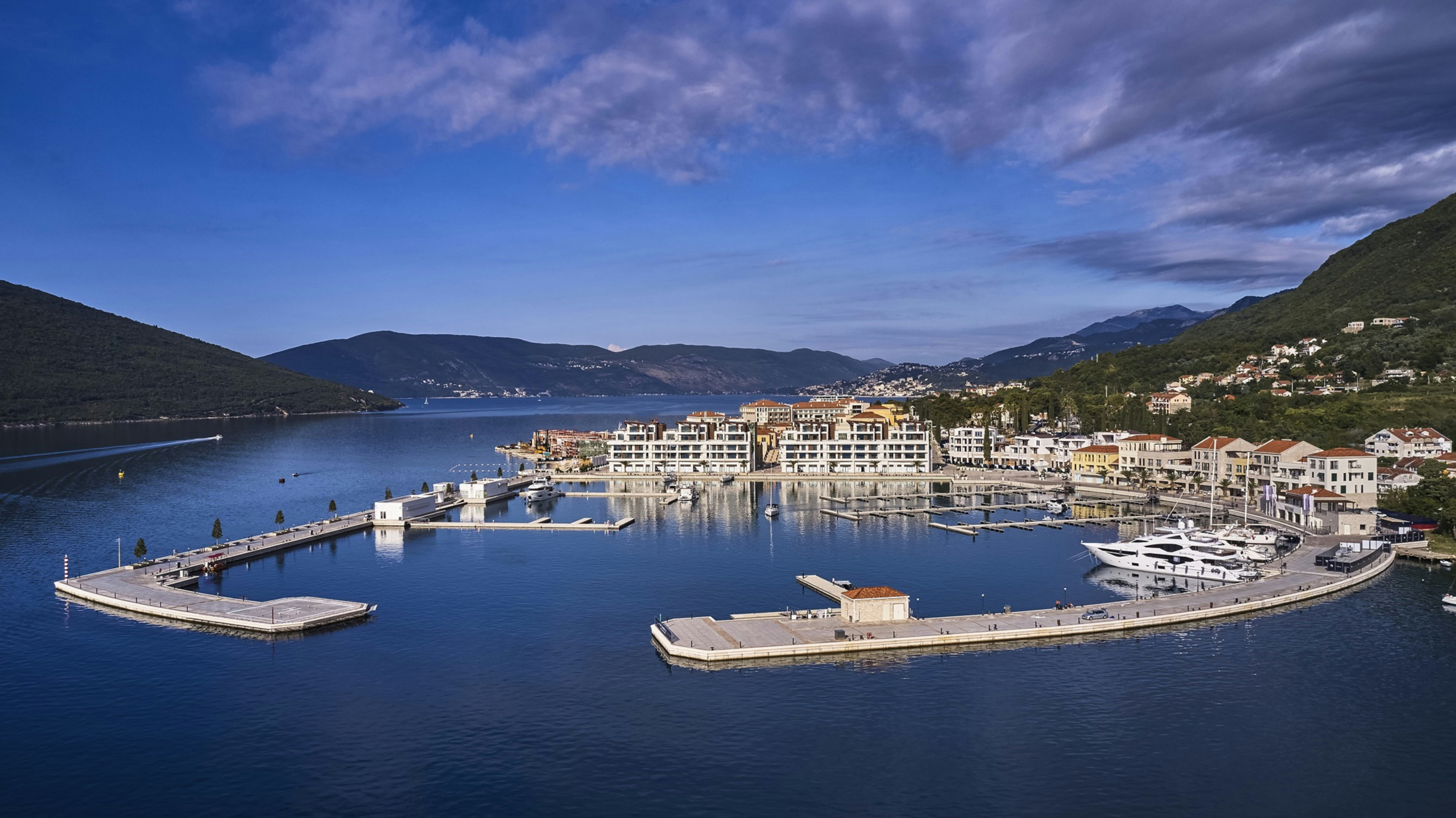 Aerial view of a coastal marina surrounded by mountains and calm blue waters under a vibrant sky.