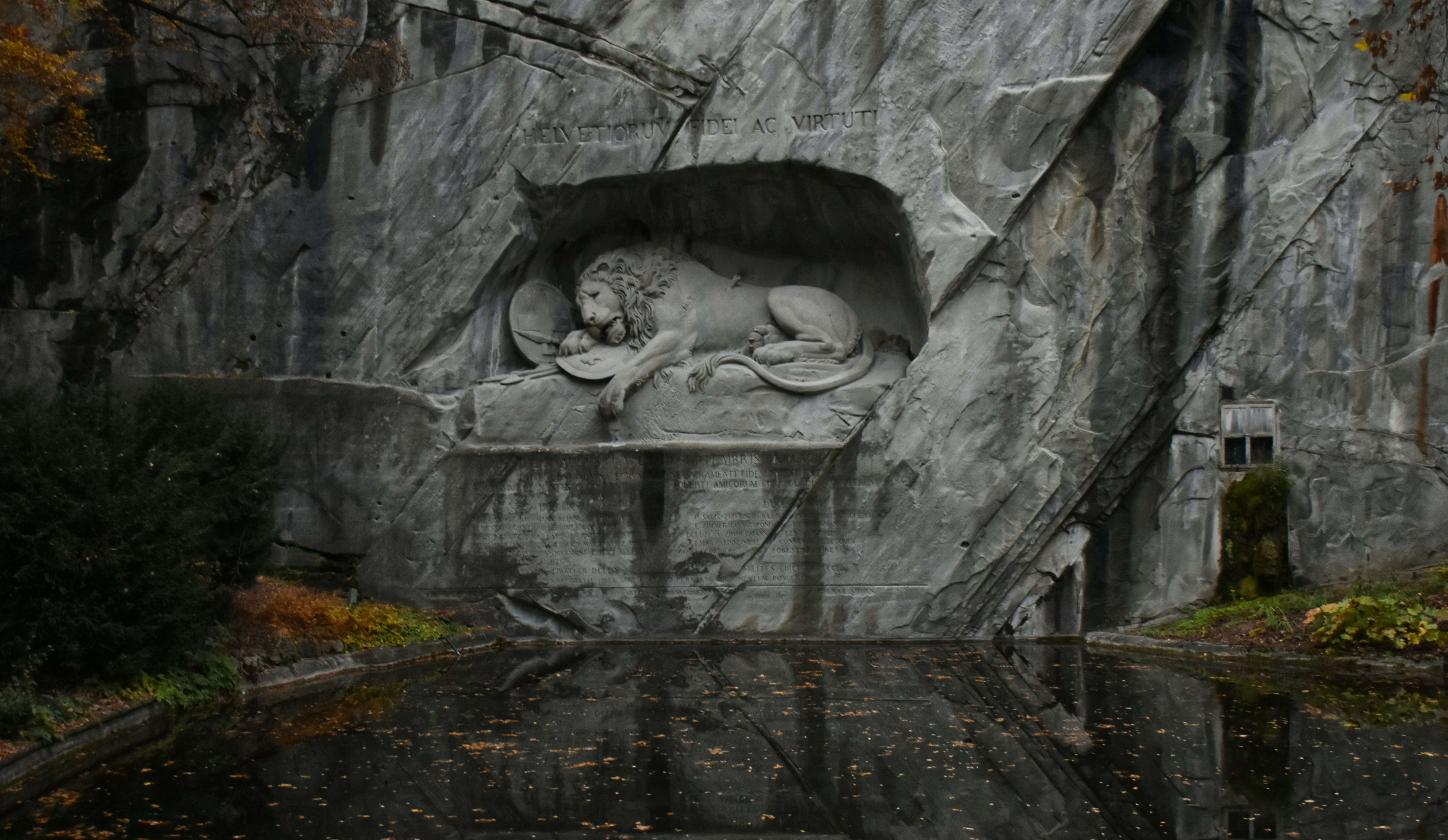 Lion sculpture carved into a rock face, reflected in a still pond, surrounded by autumn foliage.