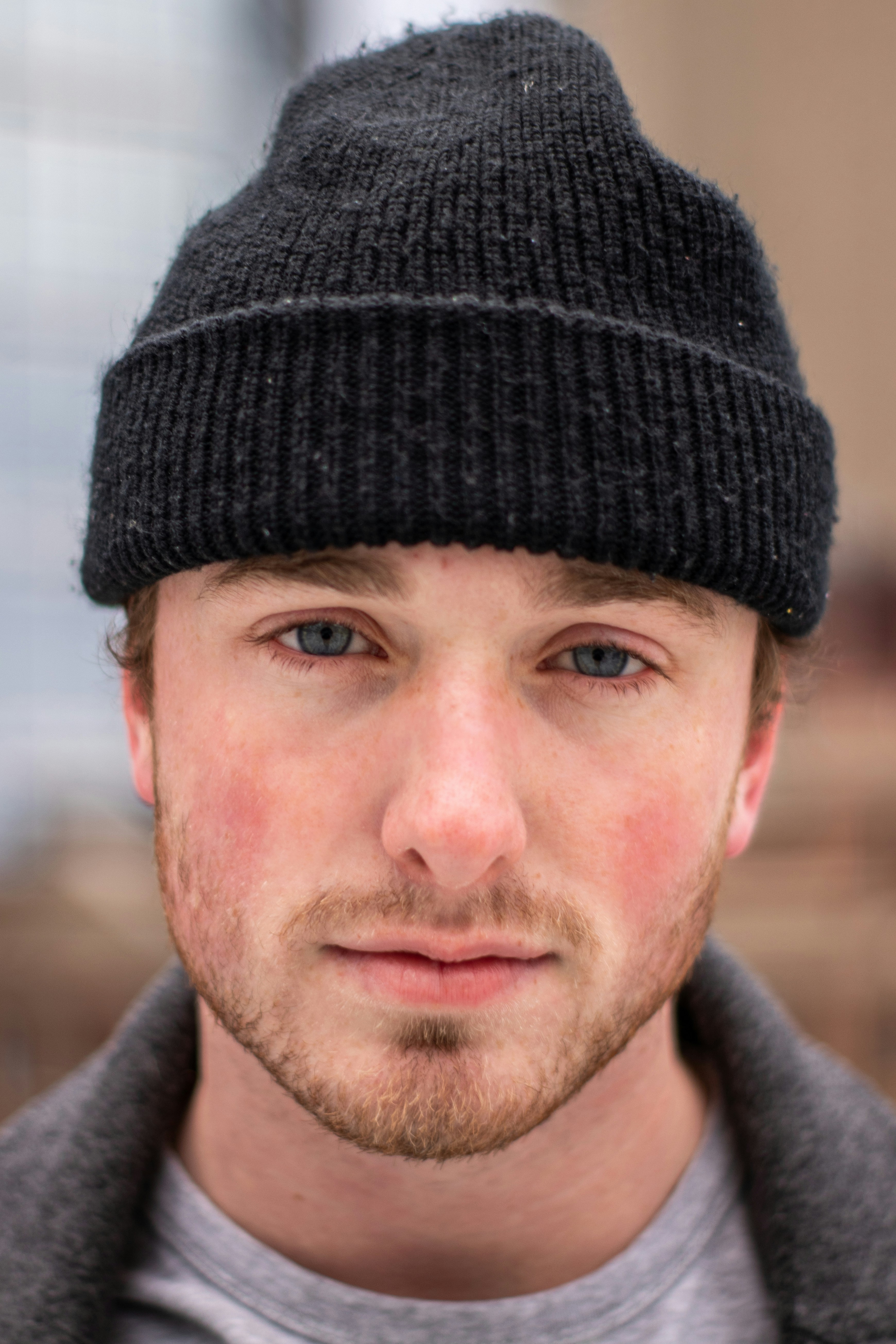 Young man with a black beanie and gray shirt gazes intently, set against a blurred urban backdrop.