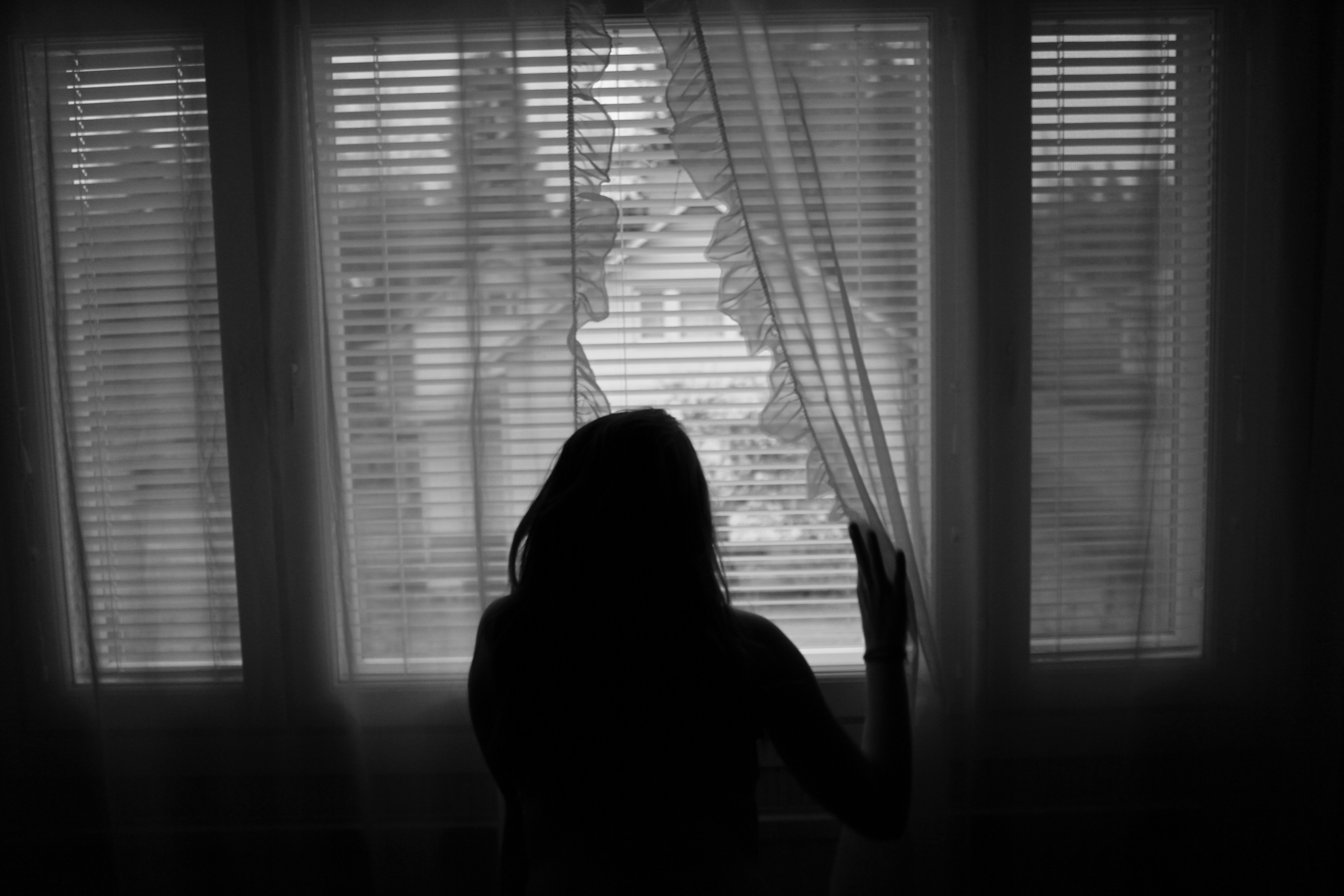 Silhouette of a figure gently pulling back a curtain, revealing a blurred view outside. The interplay of light and shadow creates a contemplative atmosphere.