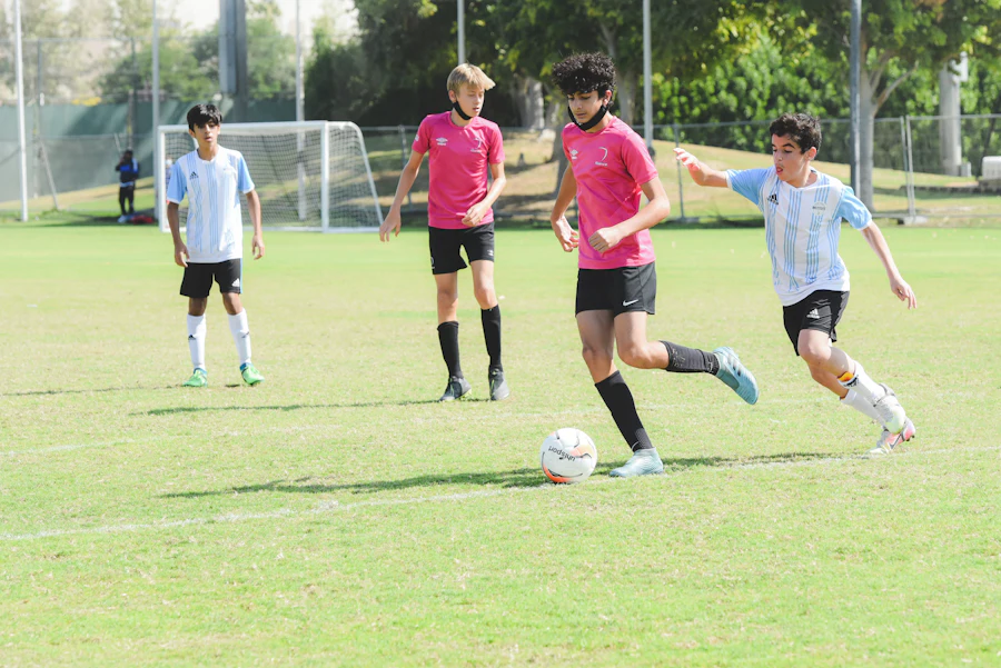 Youth soccer players on a sunlit field with a pink-jerseyed player dribbling the ball while an opponent lunges to challenge.