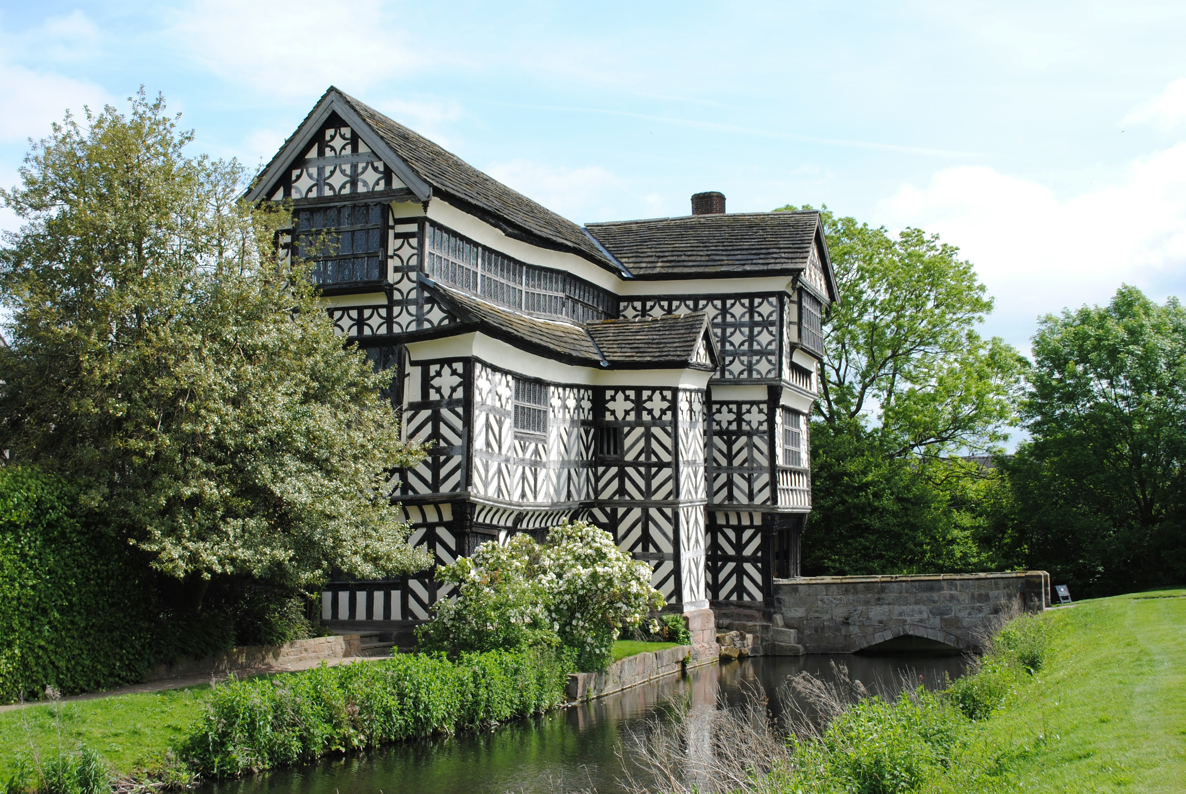 Half-timbered Tudor house with intricate designs beside a tranquil canal under a blue sky.