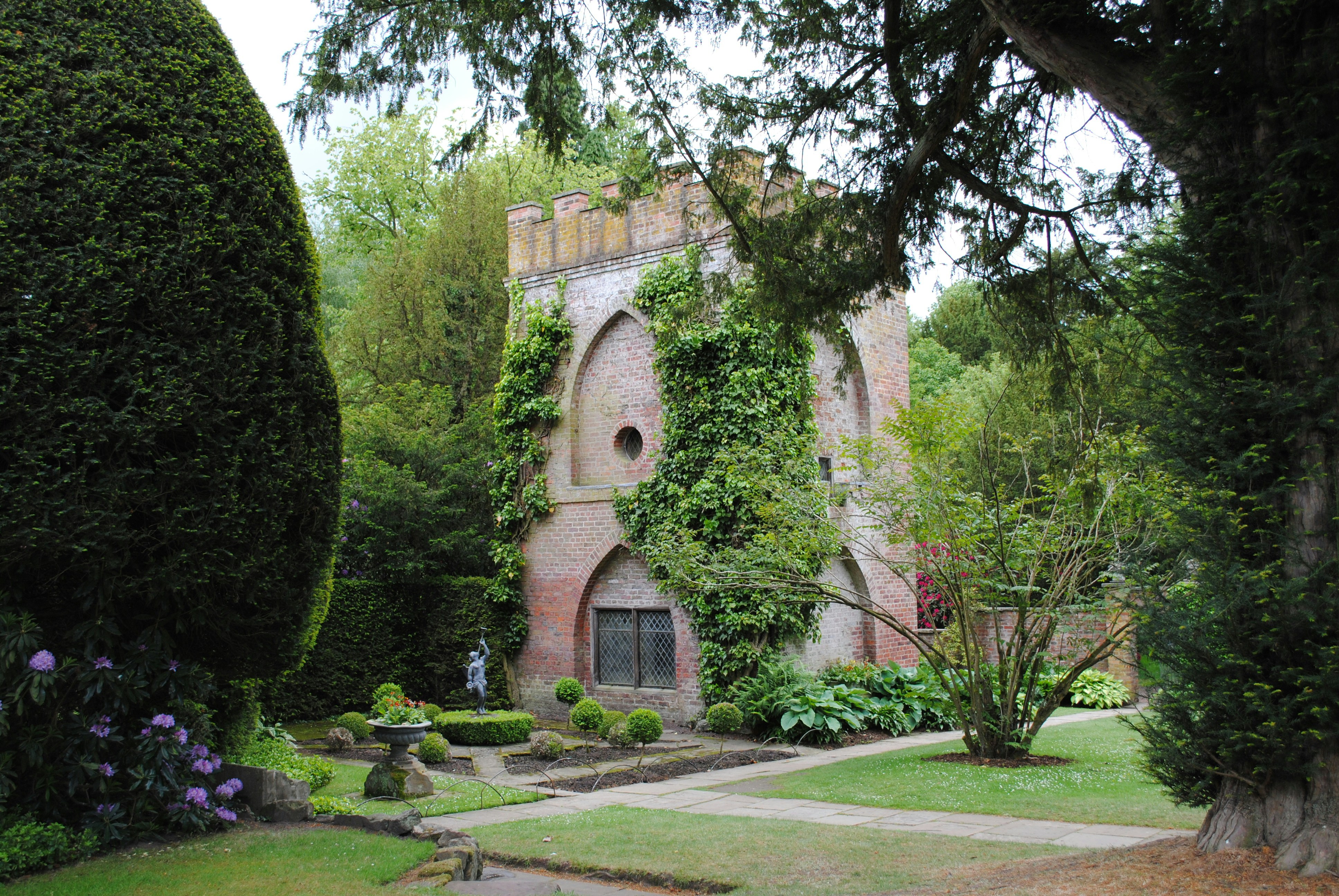 A charming brick structure entwined with greenery, surrounded by manicured gardens and ornamental plants. A serene statue adds a touch of elegance to the tranquil setting.