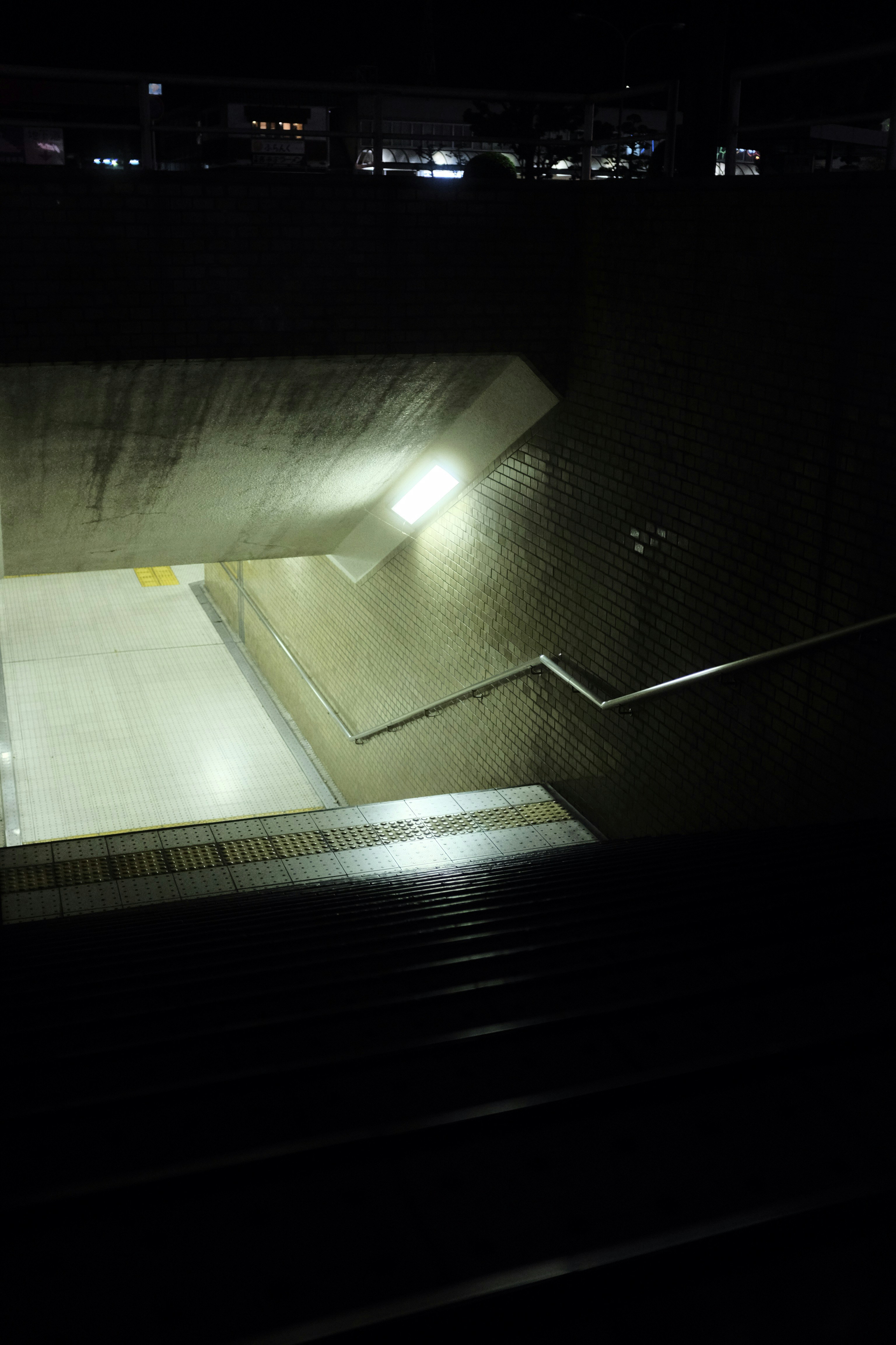 Dimly lit staircase leading into a subway entrance, with a single overhead light illuminating the concrete walls. The scene captures the essence of urban exploration.