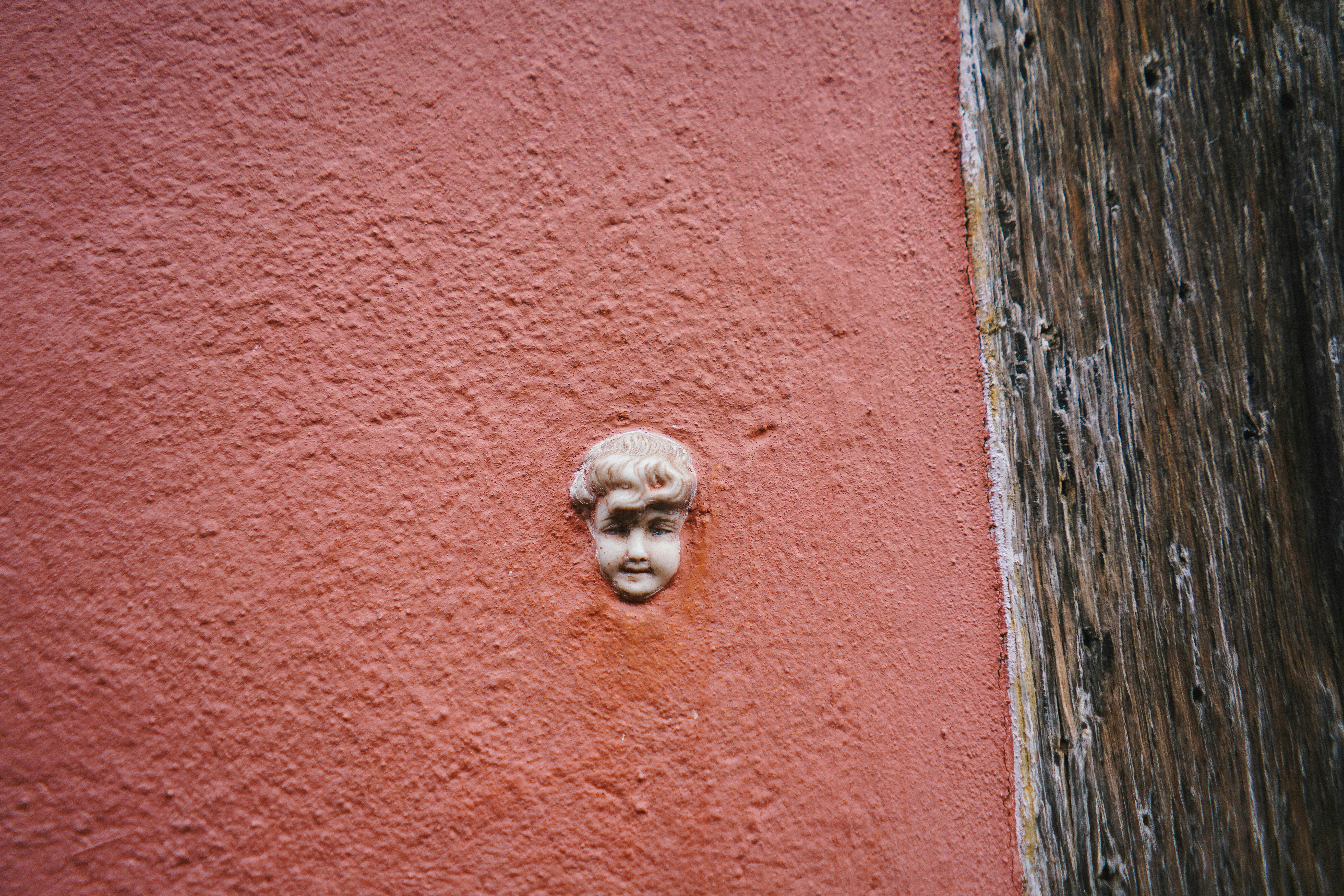 Small sculpted face embedded in a textured red wall beside a weathered wooden surface.