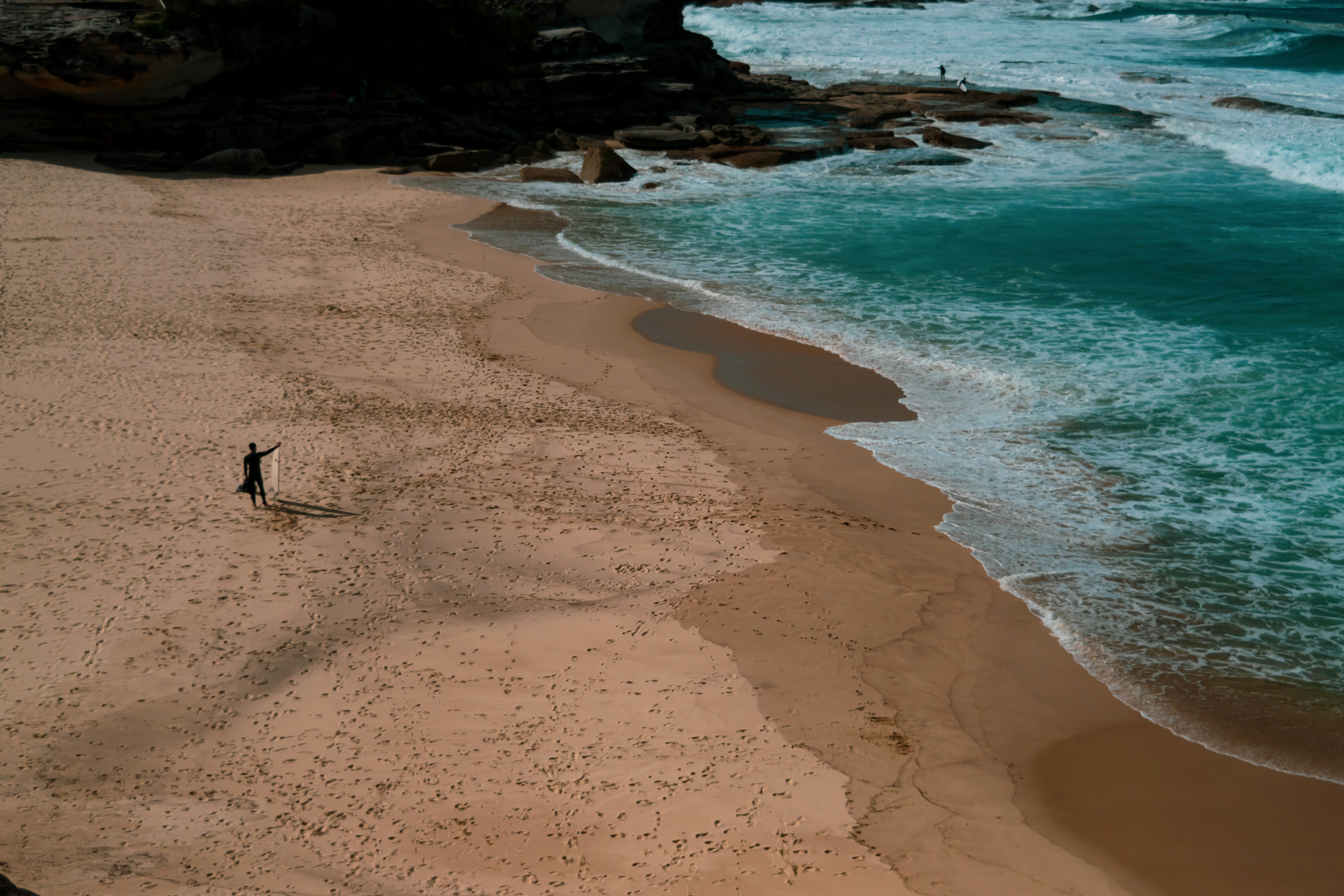 A lone figure stands on a sandy beach, casting a long shadow as waves lap at the shore. The contrasting textures of sand and water create a serene coastal scene.