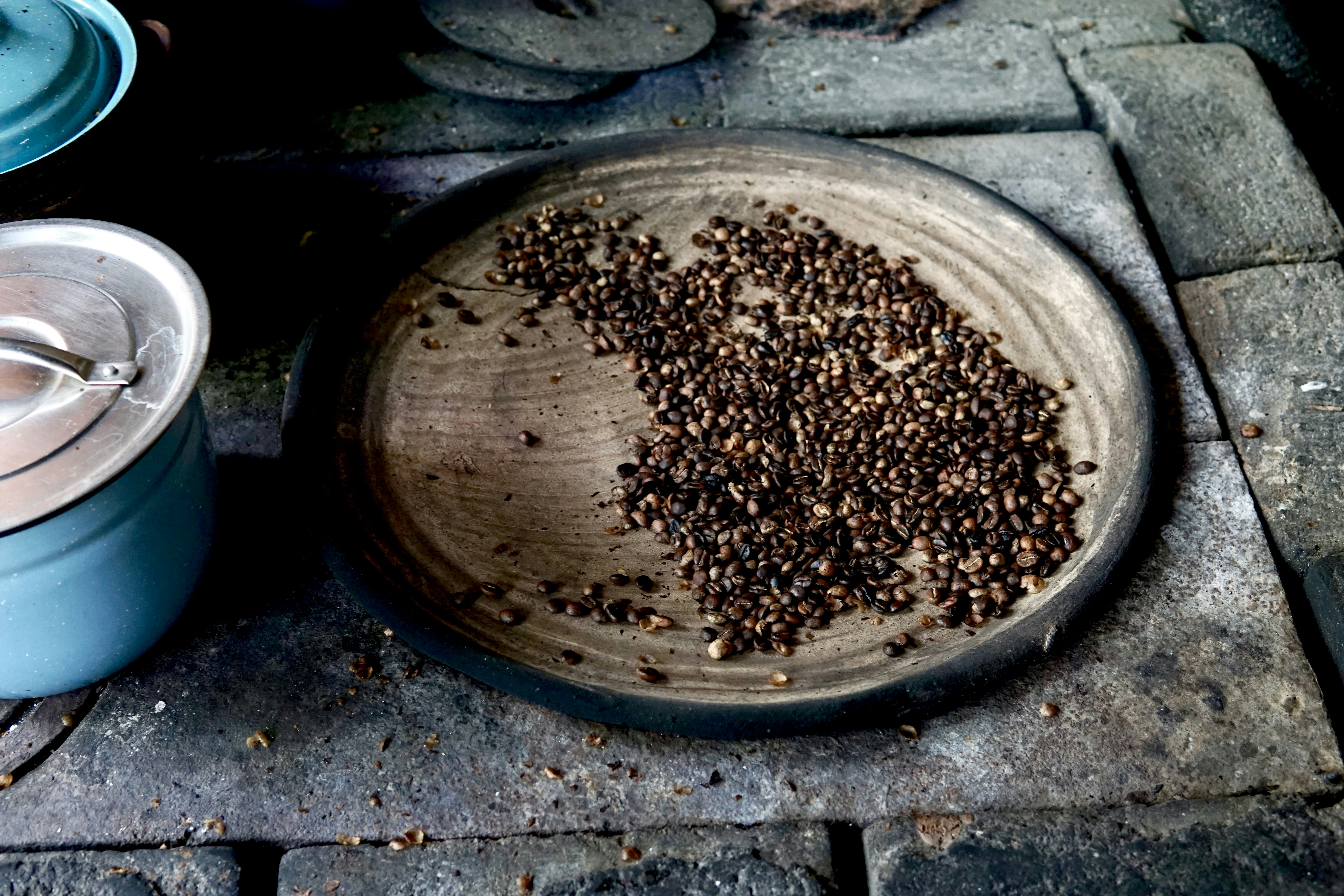Wooden tray of coffee beans resting on a stone surface beside a metal pot.
