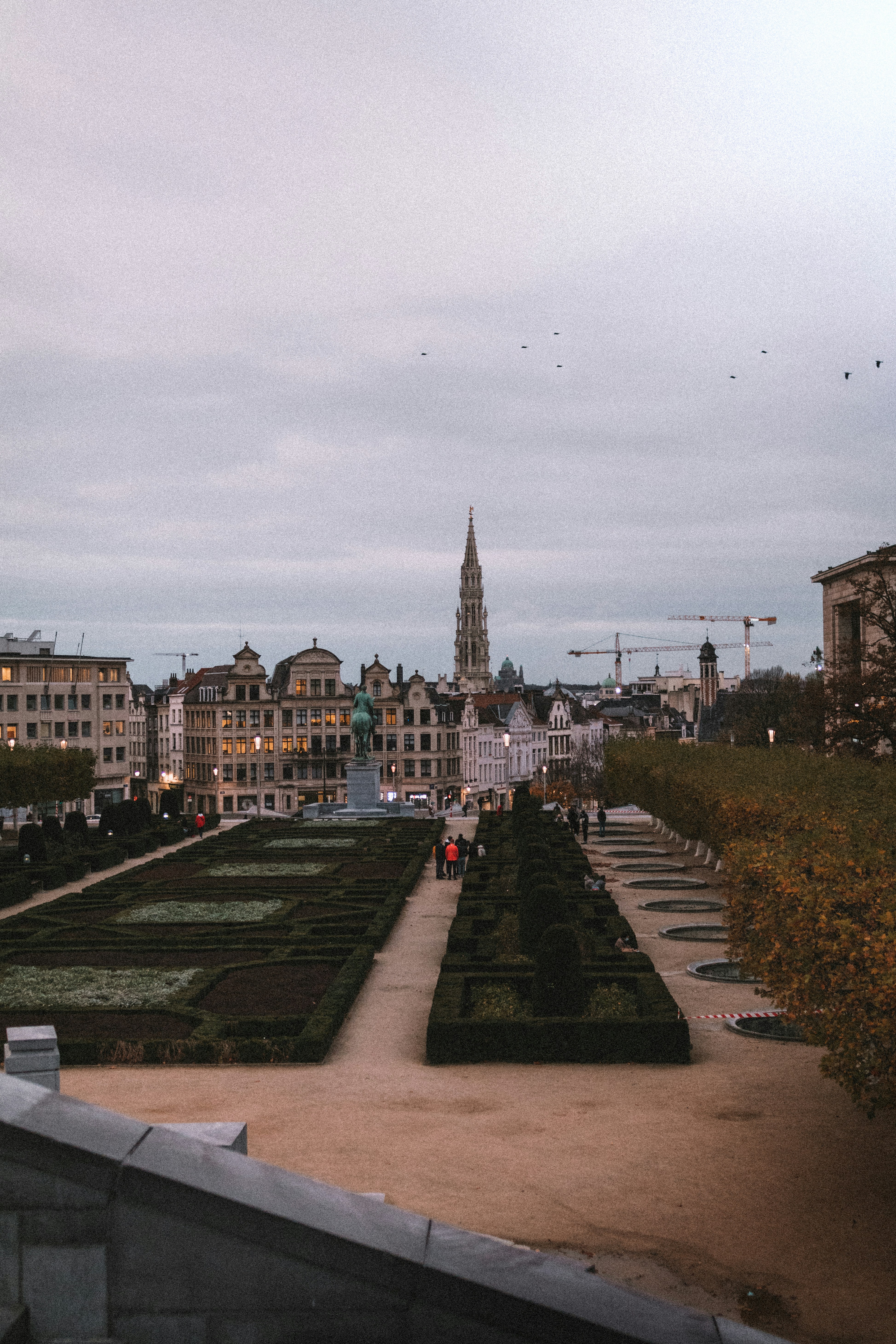 Elegant gardens lined with hedges and pathways, showcasing the cityscape of Brussels under a twilight sky.