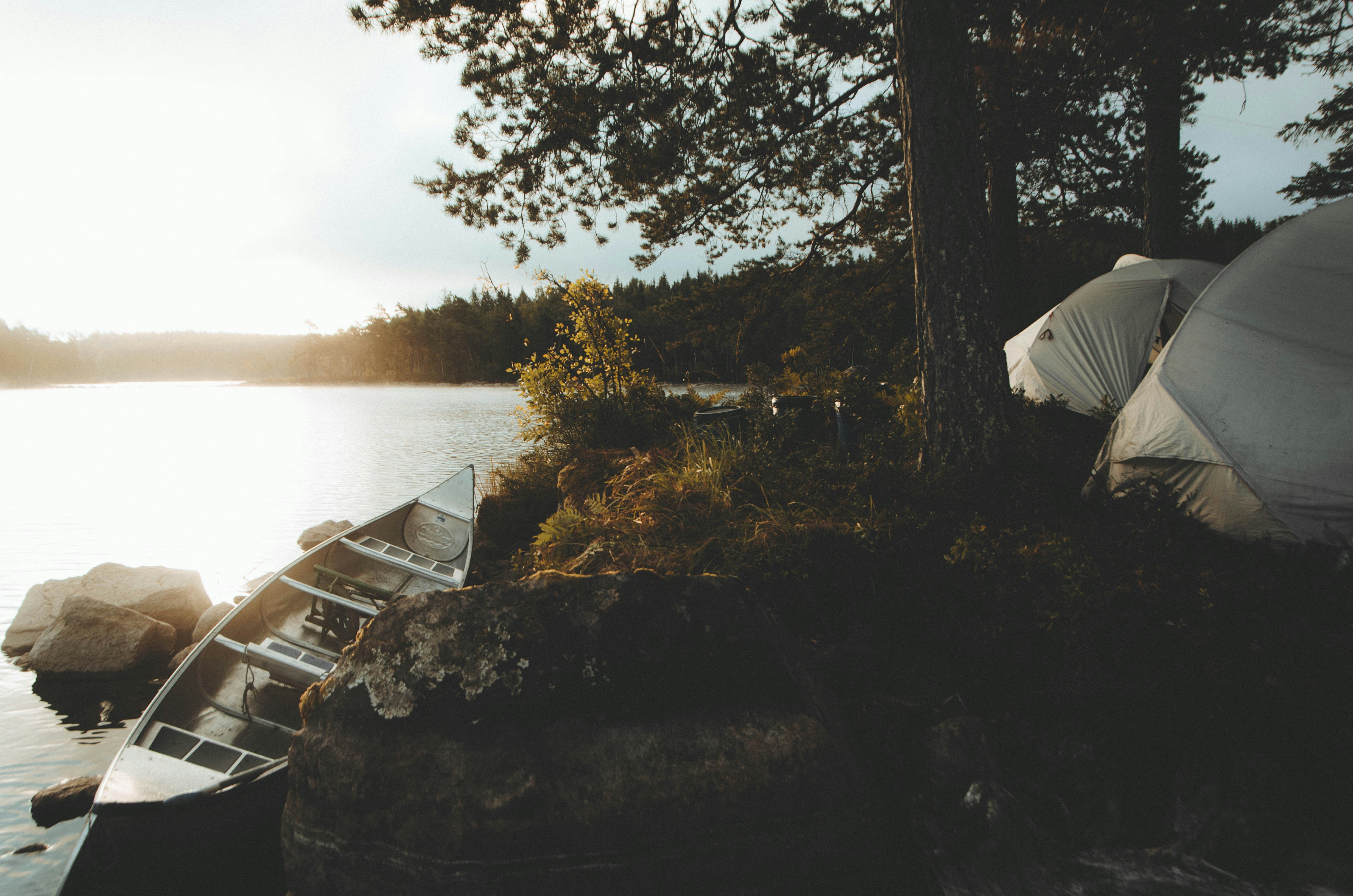 Canoe resting on rocky shore with a tent nearby, illuminated by soft evening light reflecting off the tranquil lake.