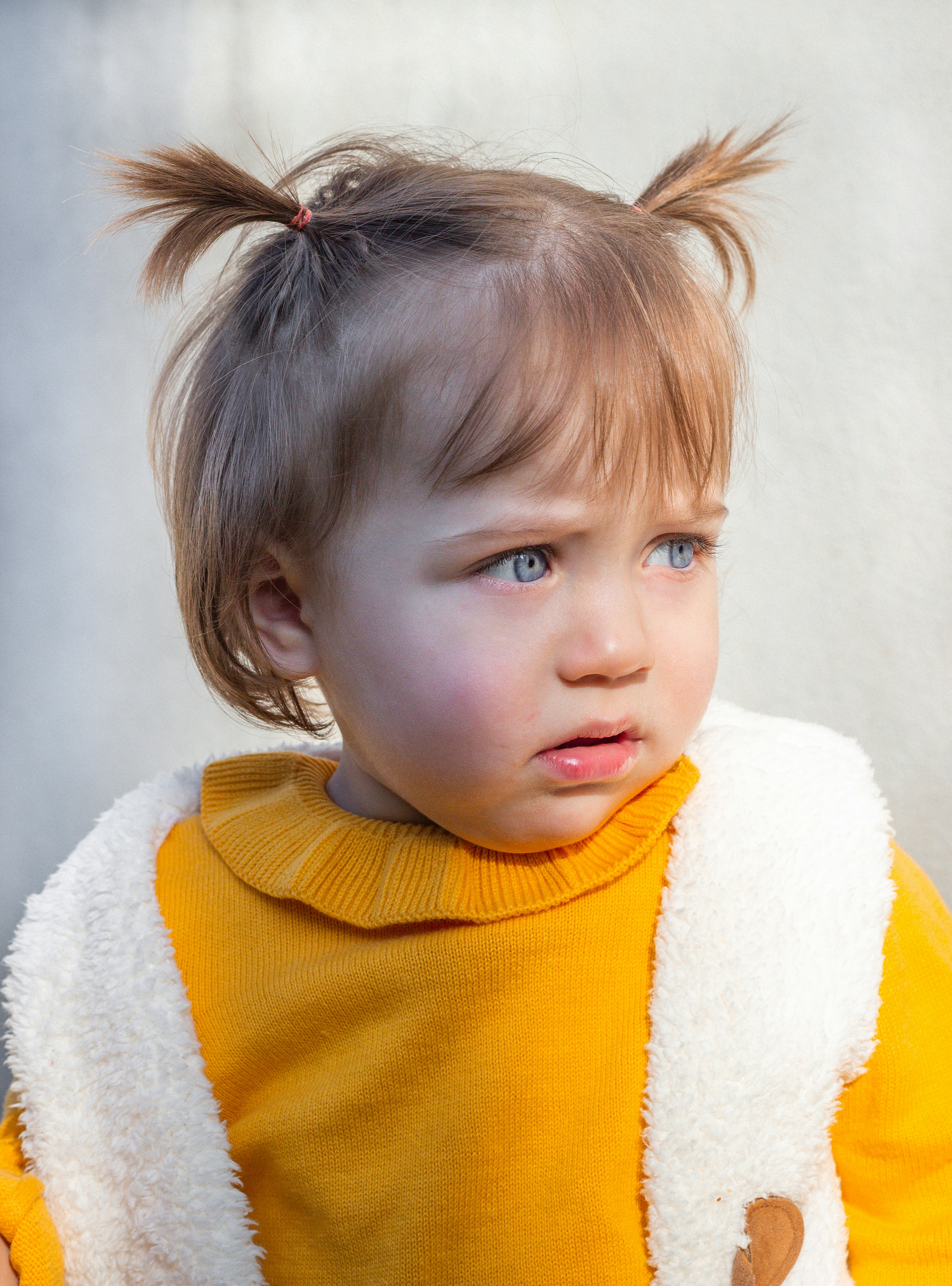 Young child with pigtails wearing a bright yellow sweater and a fluffy vest, gazing thoughtfully off-camera against a soft backdrop.