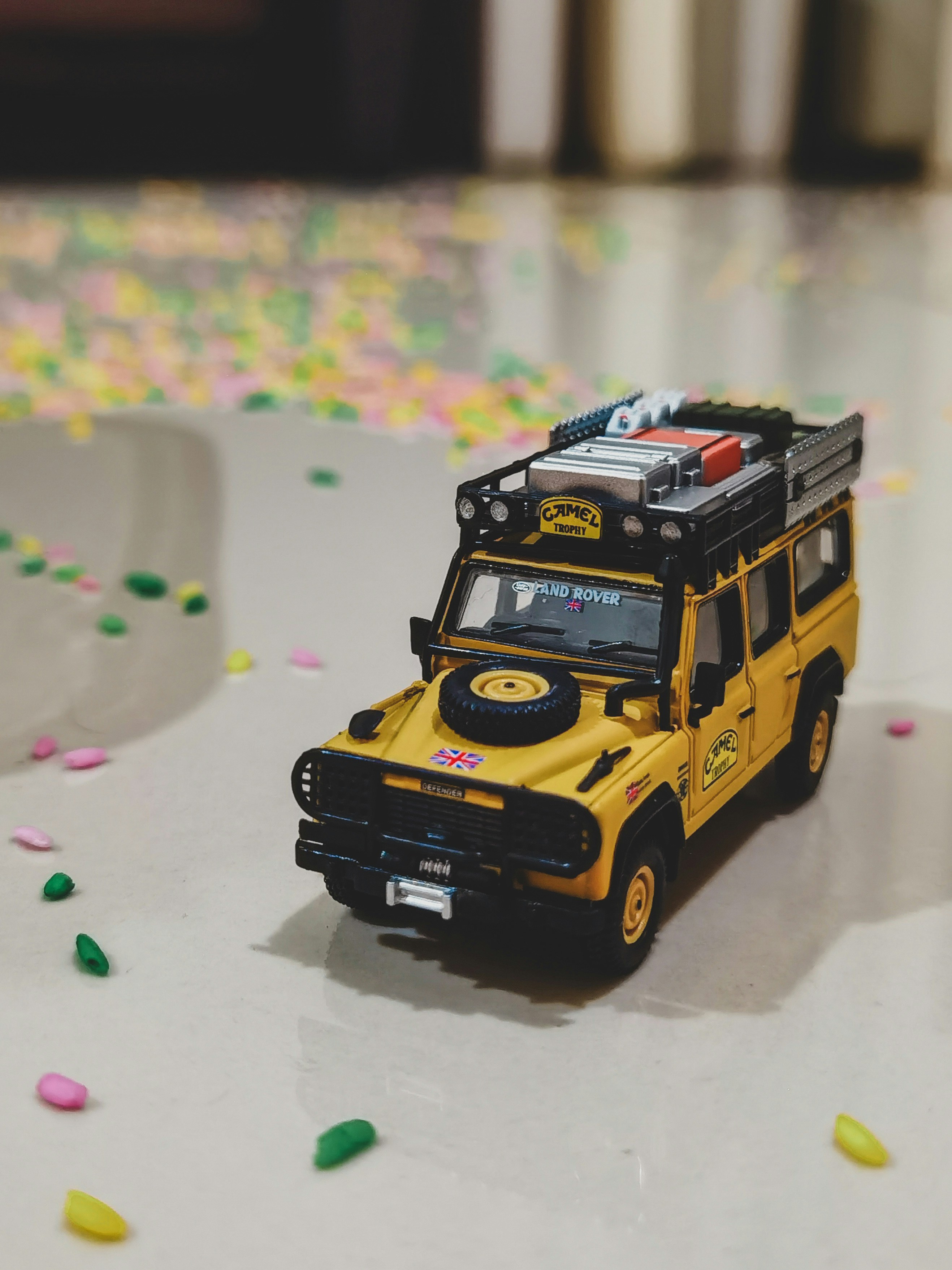 A detailed model of a yellow Land Rover, adorned with Camel Trophy branding, set against a colorful backdrop of scattered confetti.