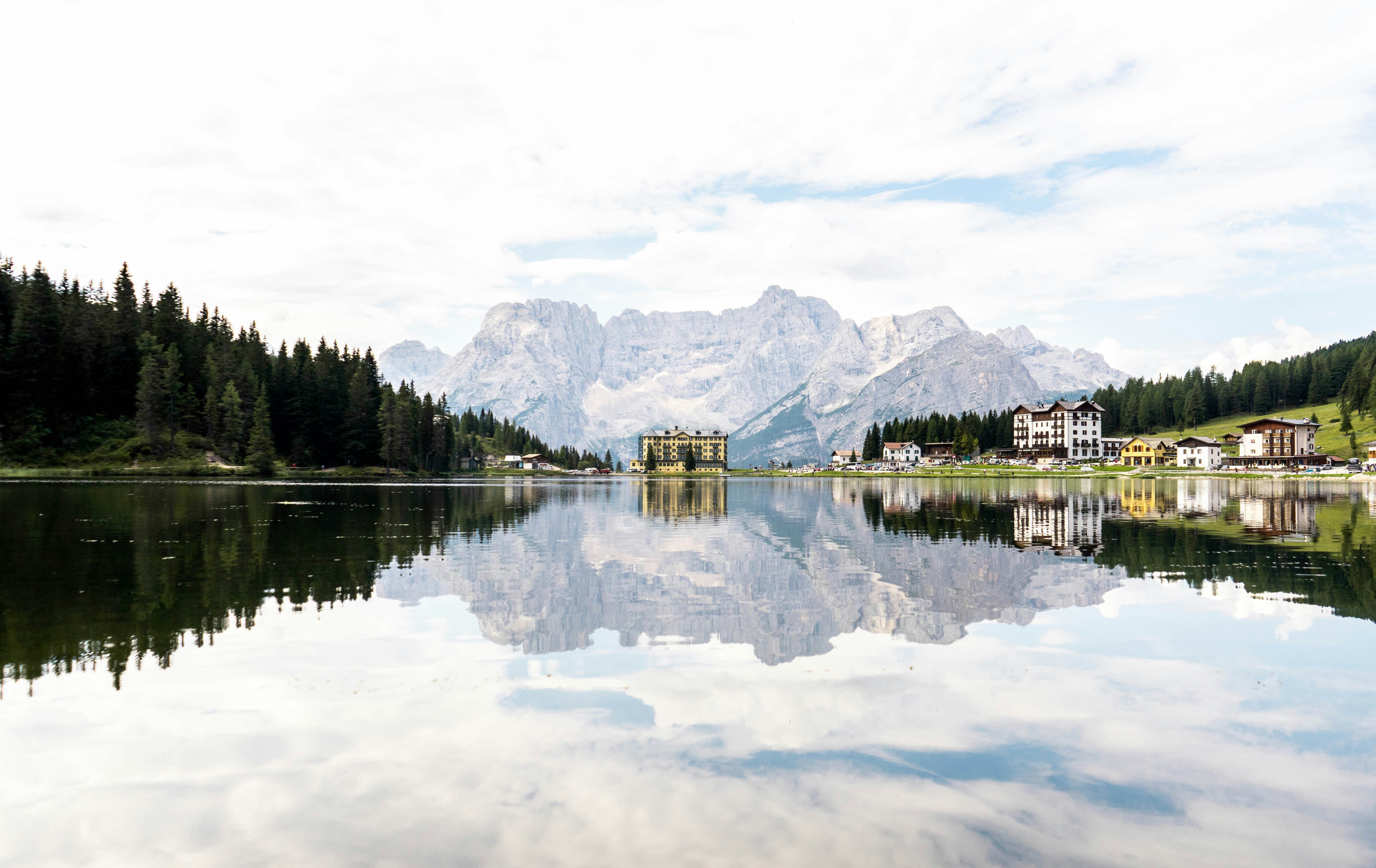 Mountain range mirrored perfectly in a tranquil lake with surrounding forest and buildings under a partly cloudy sky.