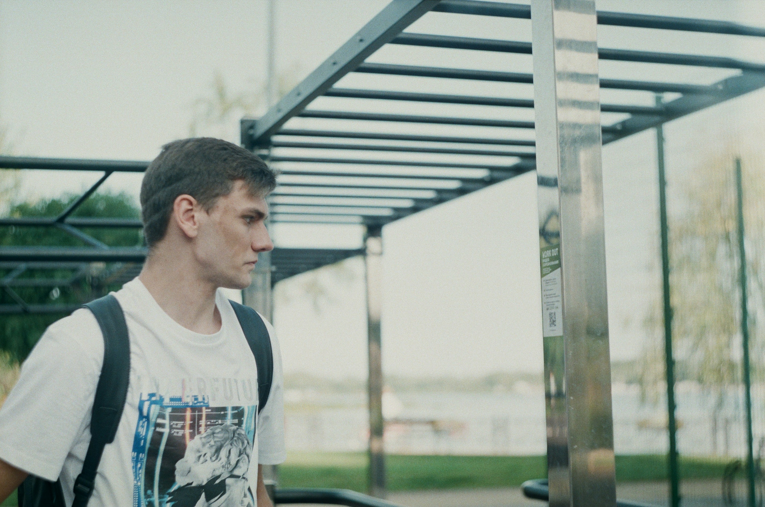 Photograph of a young man with a backpack under a steel pergola at a park, looking to the left. The image captures a candid outdoor portrait.