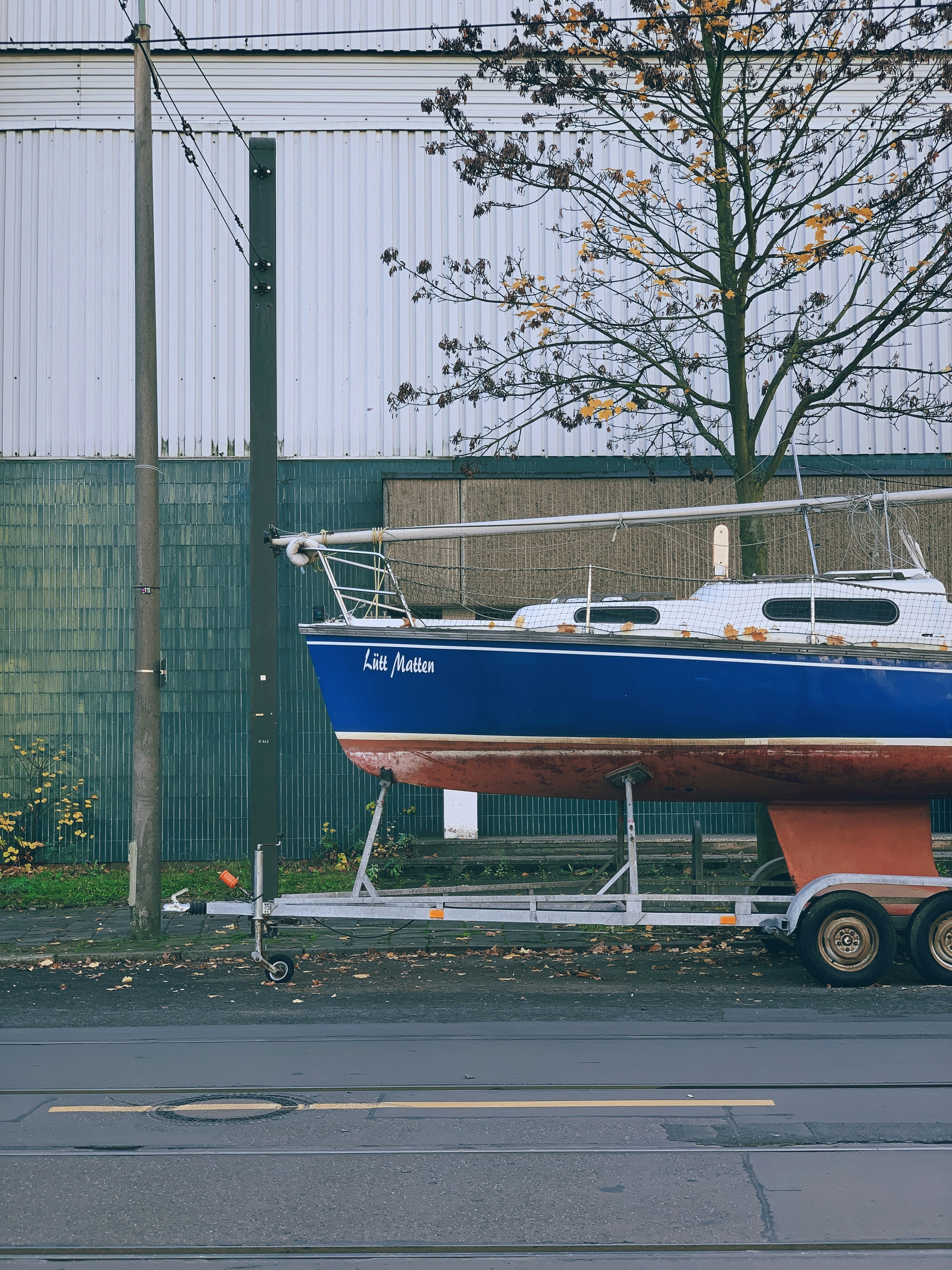 Blue Sailboat on a Street-Side Trailer: Urban Juxtaposition by a Corrugated Wall