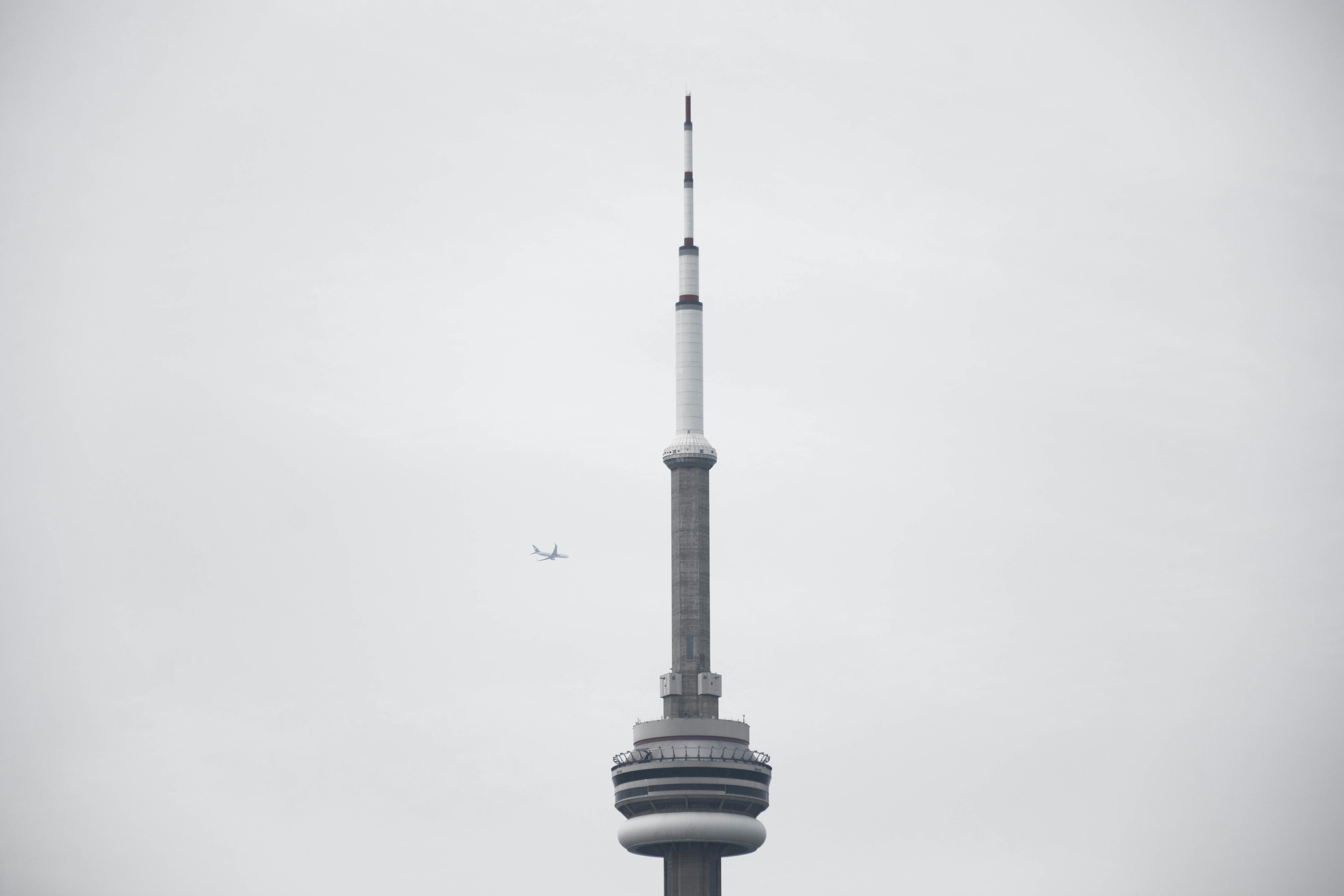 The CN Tower reaches skyward, with a small airplane flying in the distance against a cloudy backdrop.