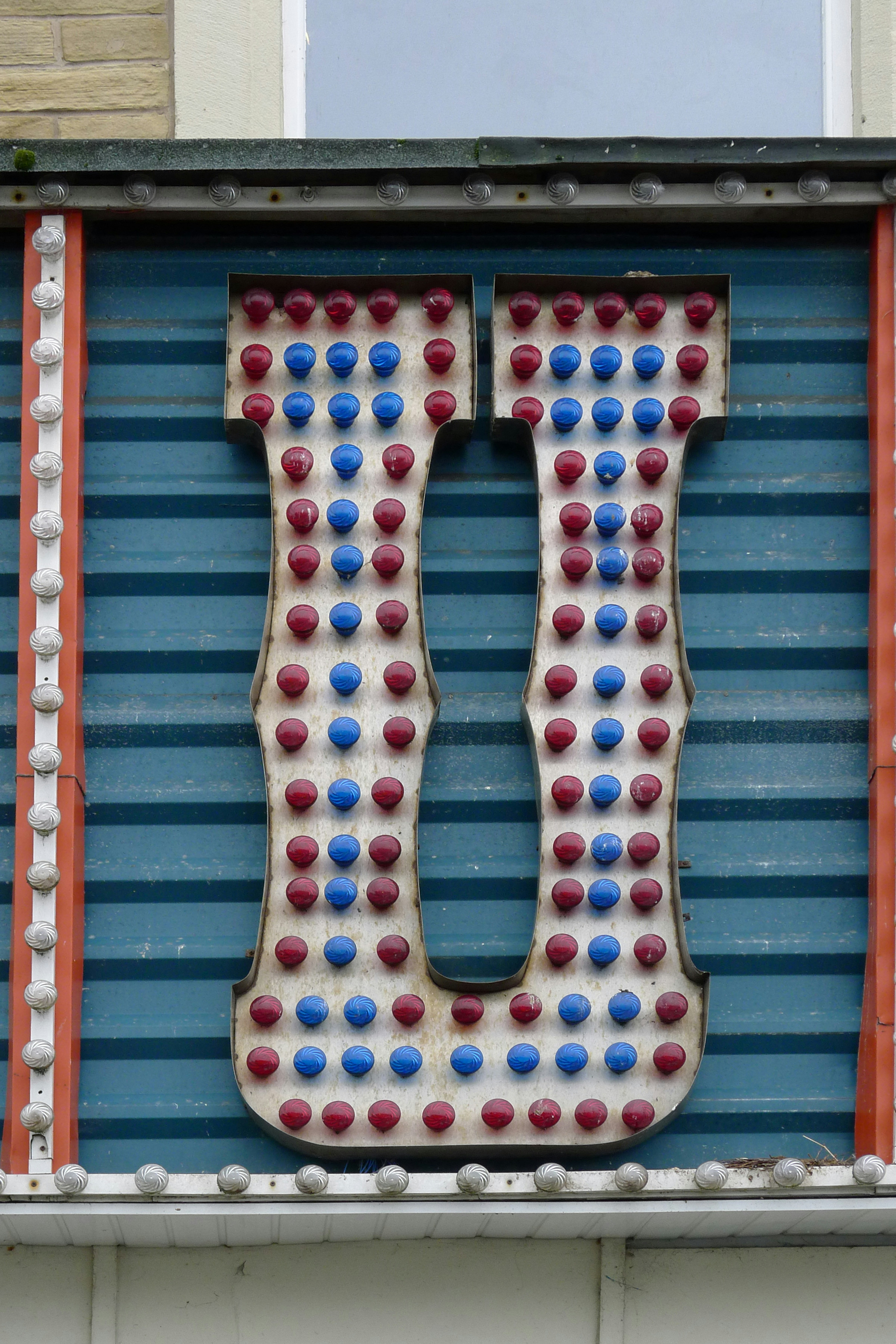 Photograph of a large U-shaped marquee sign studded with red and blue bulbs against a blue corrugated metal backdrop.