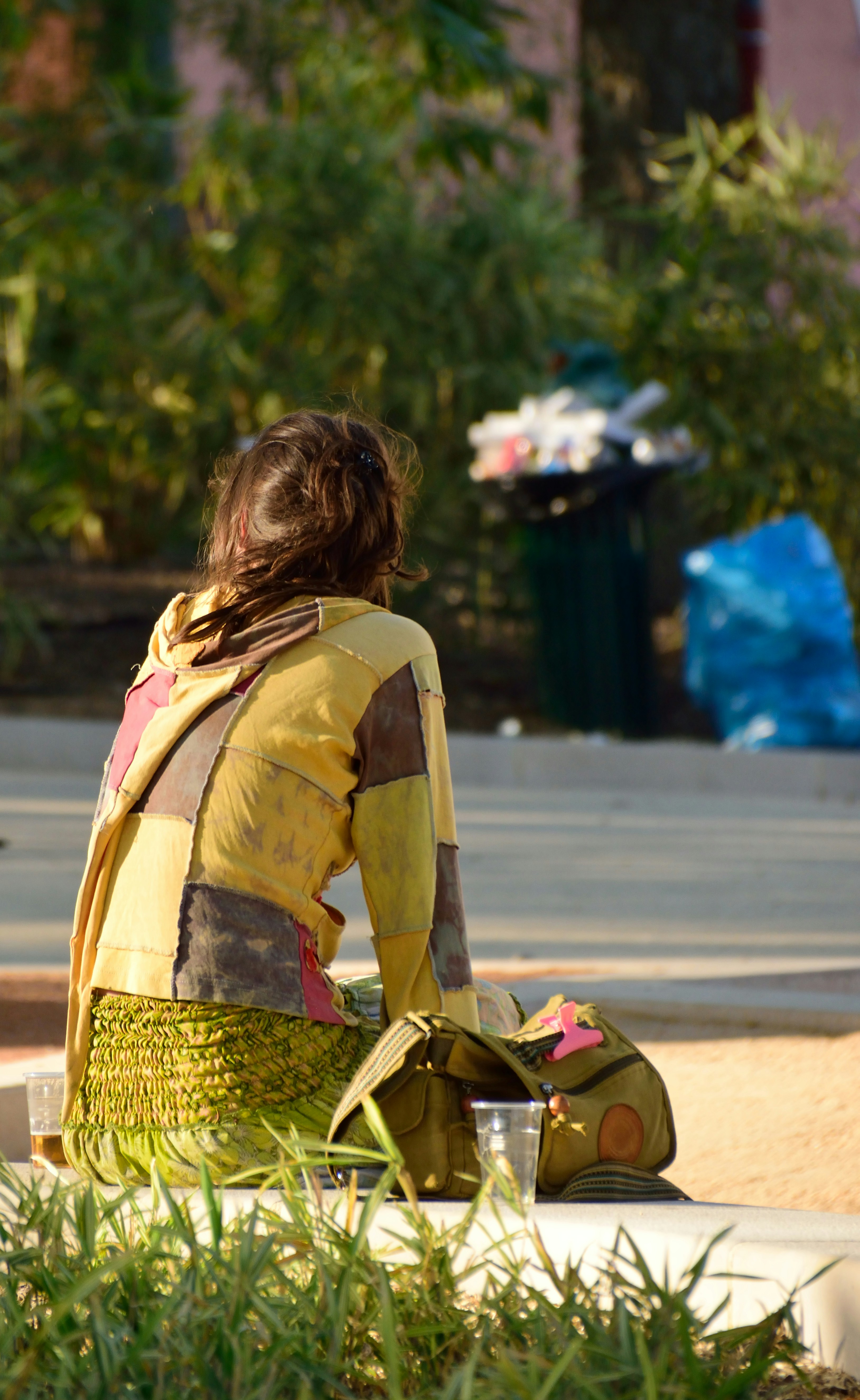 Person in a colorful patchwork jacket sitting on a bench with a green bag beside them.