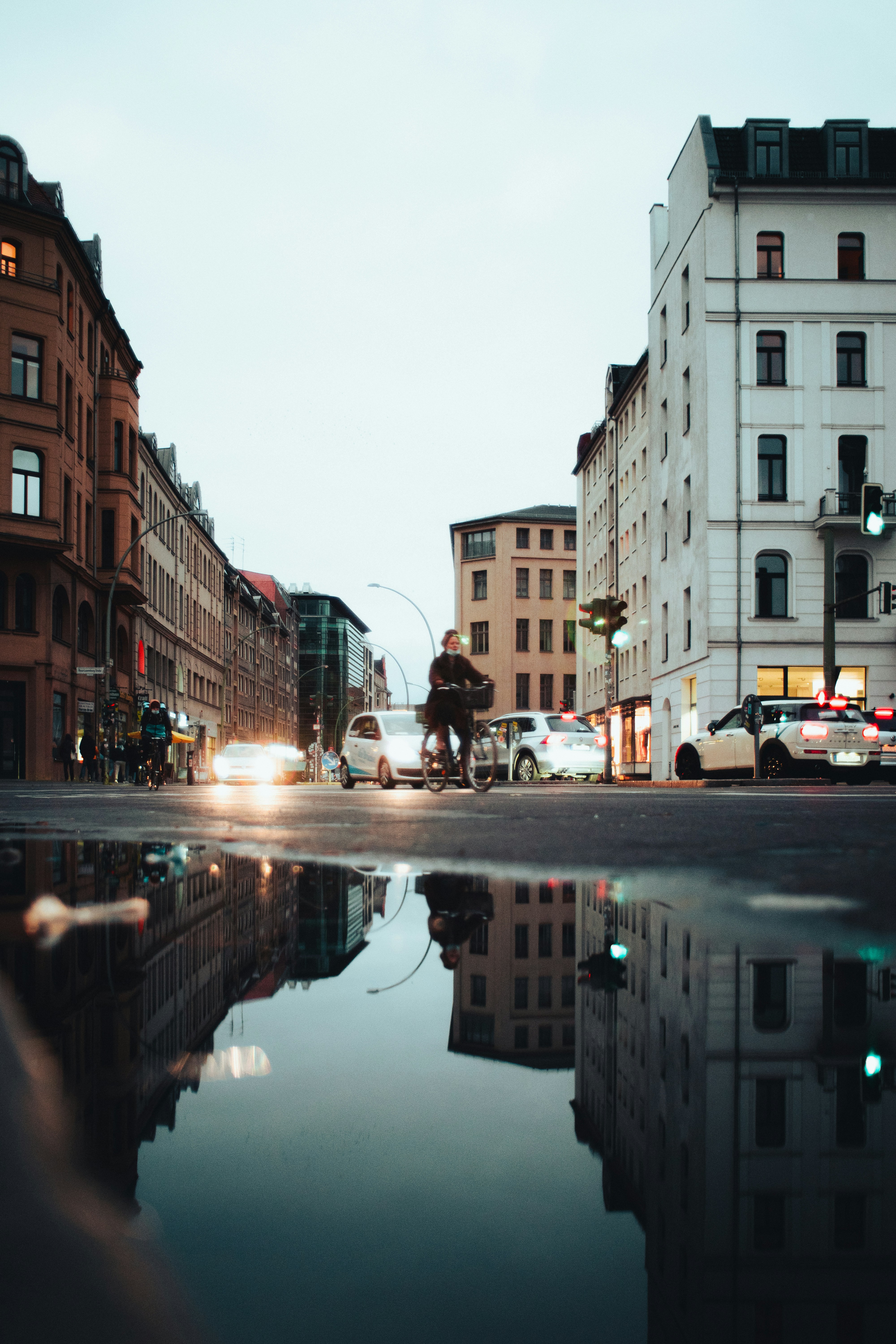 A cyclist navigates a city street, with buildings mirrored in a puddle, capturing the essence of urban movement and architecture.