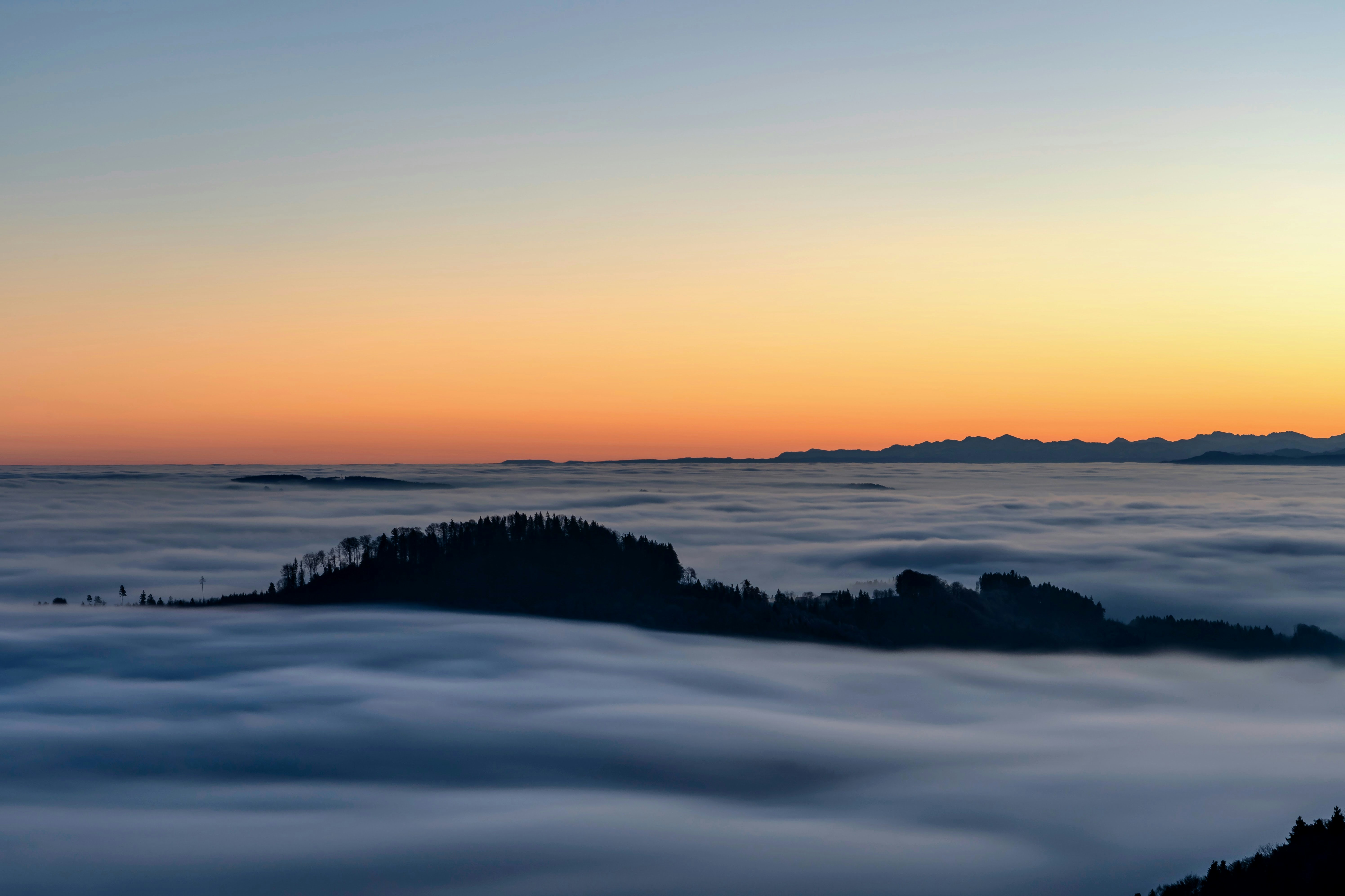 A serene landscape showcasing rolling clouds beneath a tranquil sunrise, with silhouettes of distant mountains on the horizon.