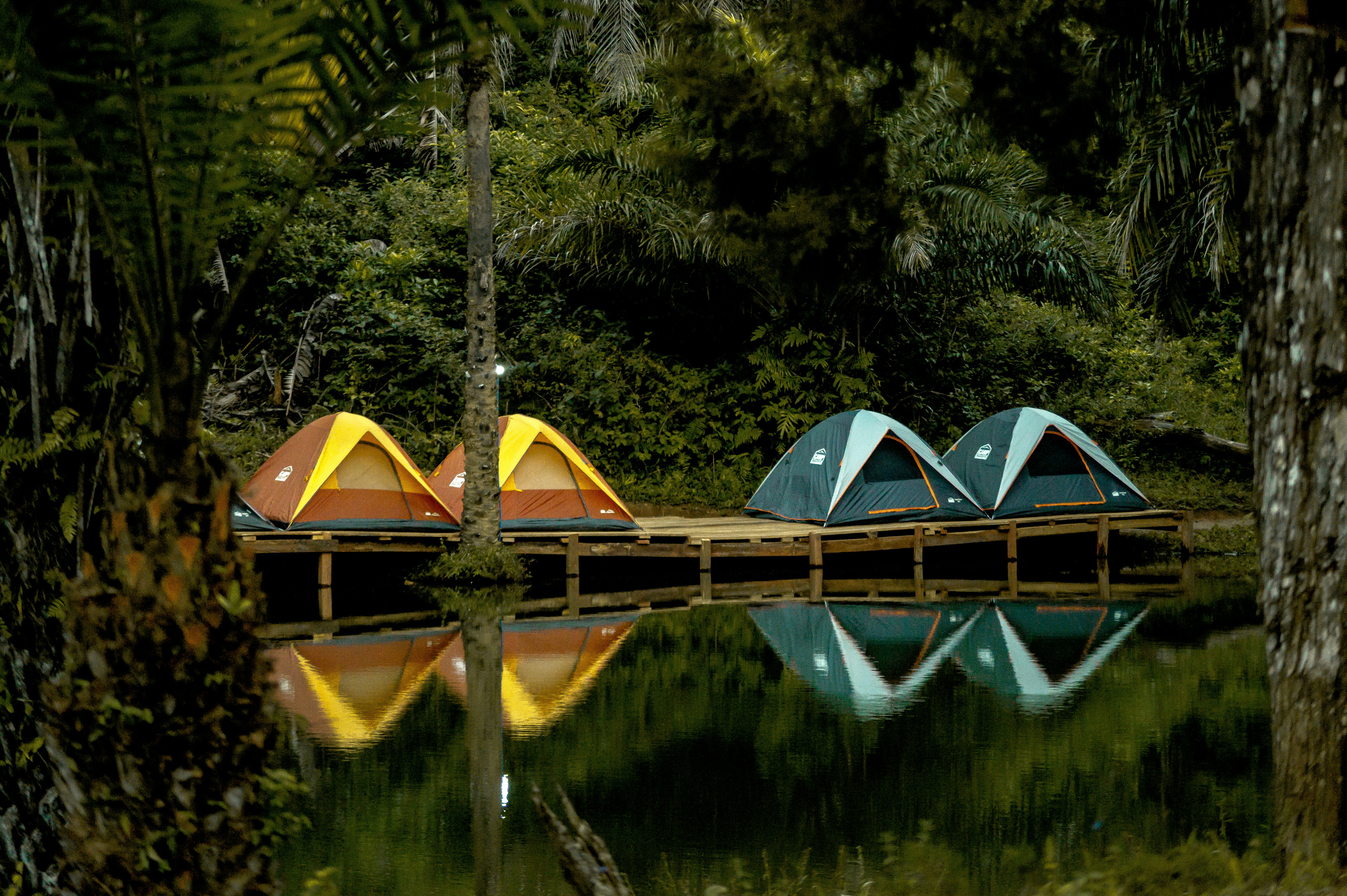 Colorful tents arranged on a wooden platform by a tranquil lake, surrounded by lush greenery. The still water perfectly mirrors the vibrant tents.