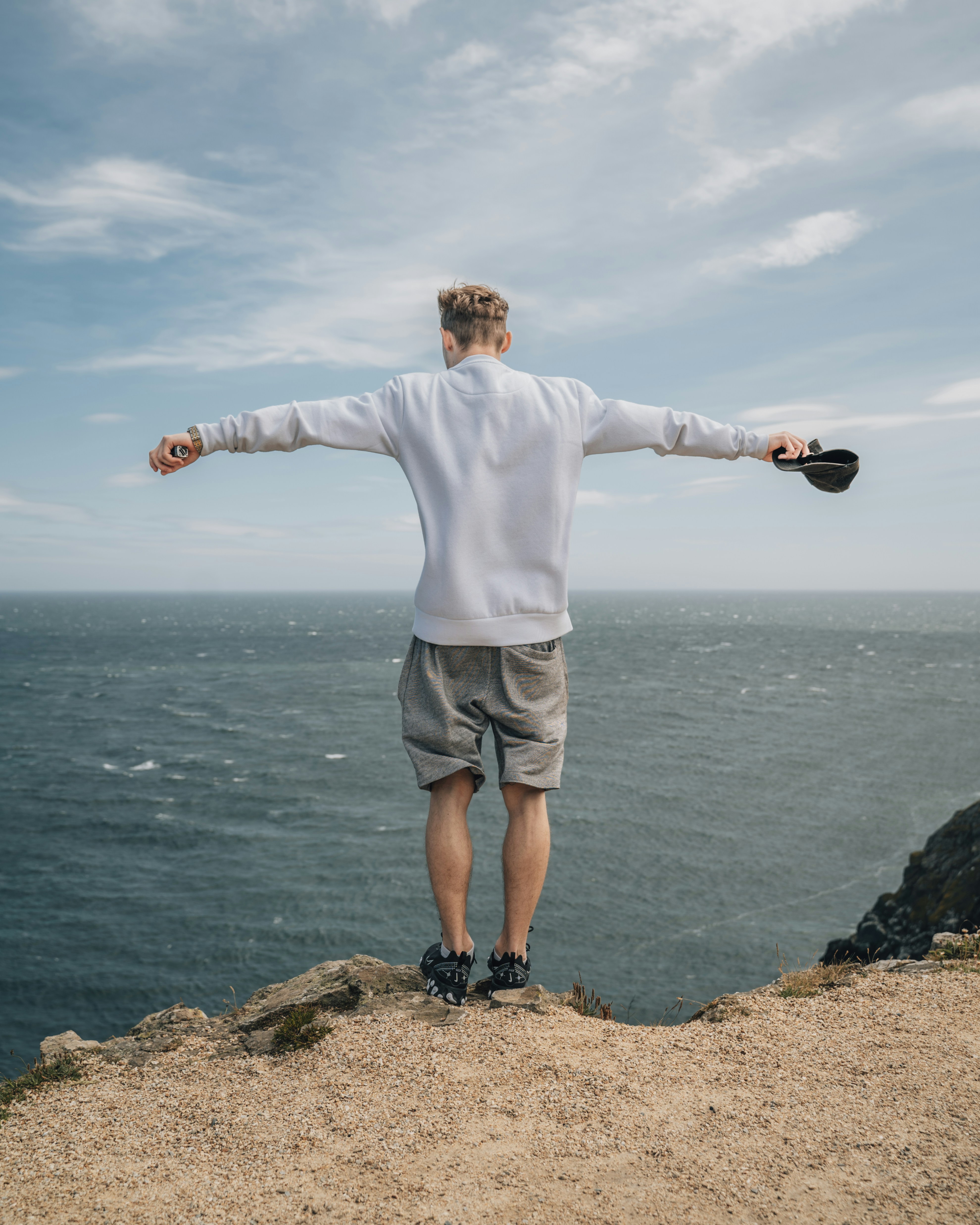 Individual standing on a cliff's edge, arms outstretched, overlooking a vast ocean under a cloudy sky.