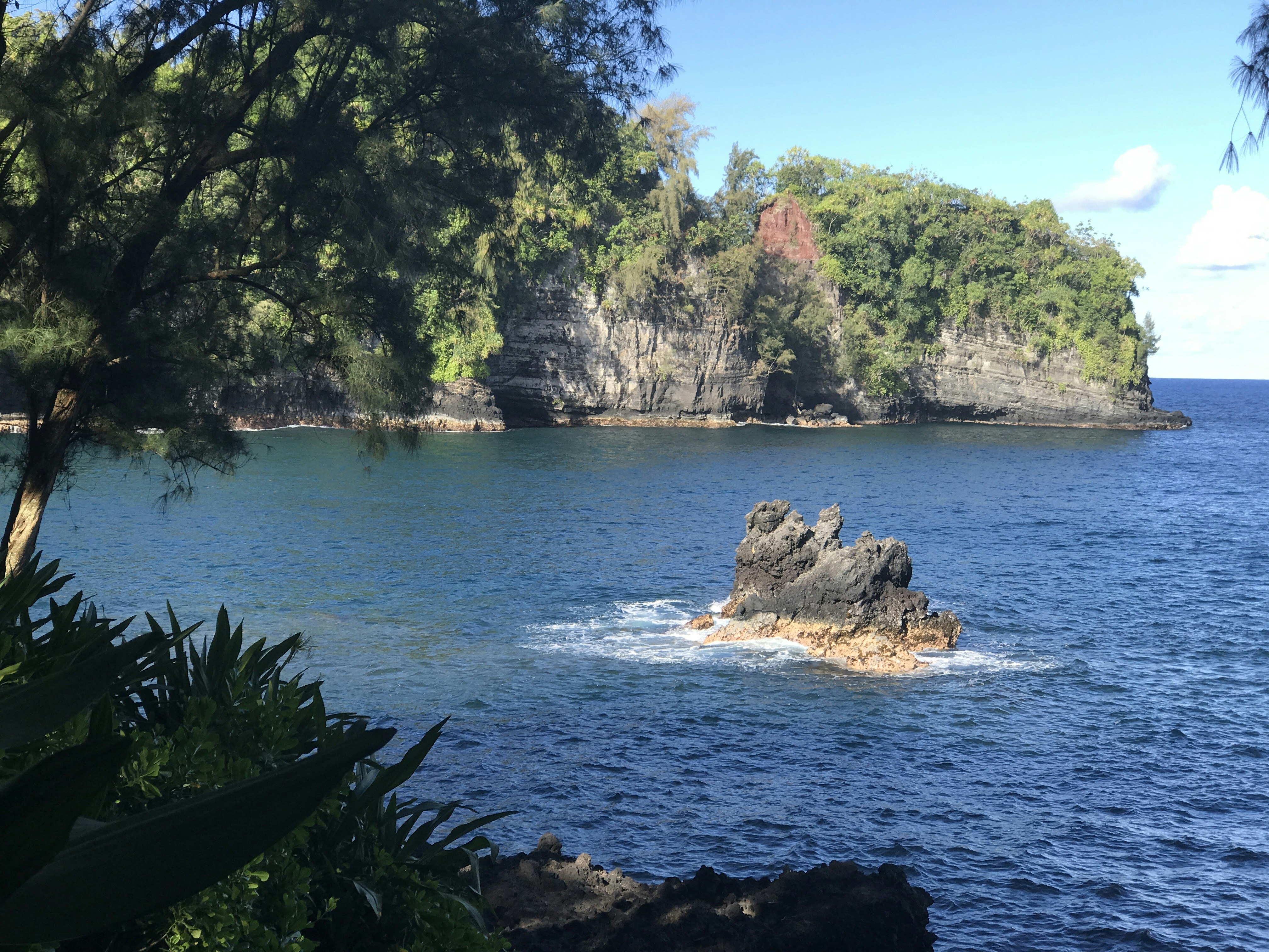 Rock formation emerging from tranquil waters, surrounded by lush greenery and rugged cliffs under a clear blue sky.