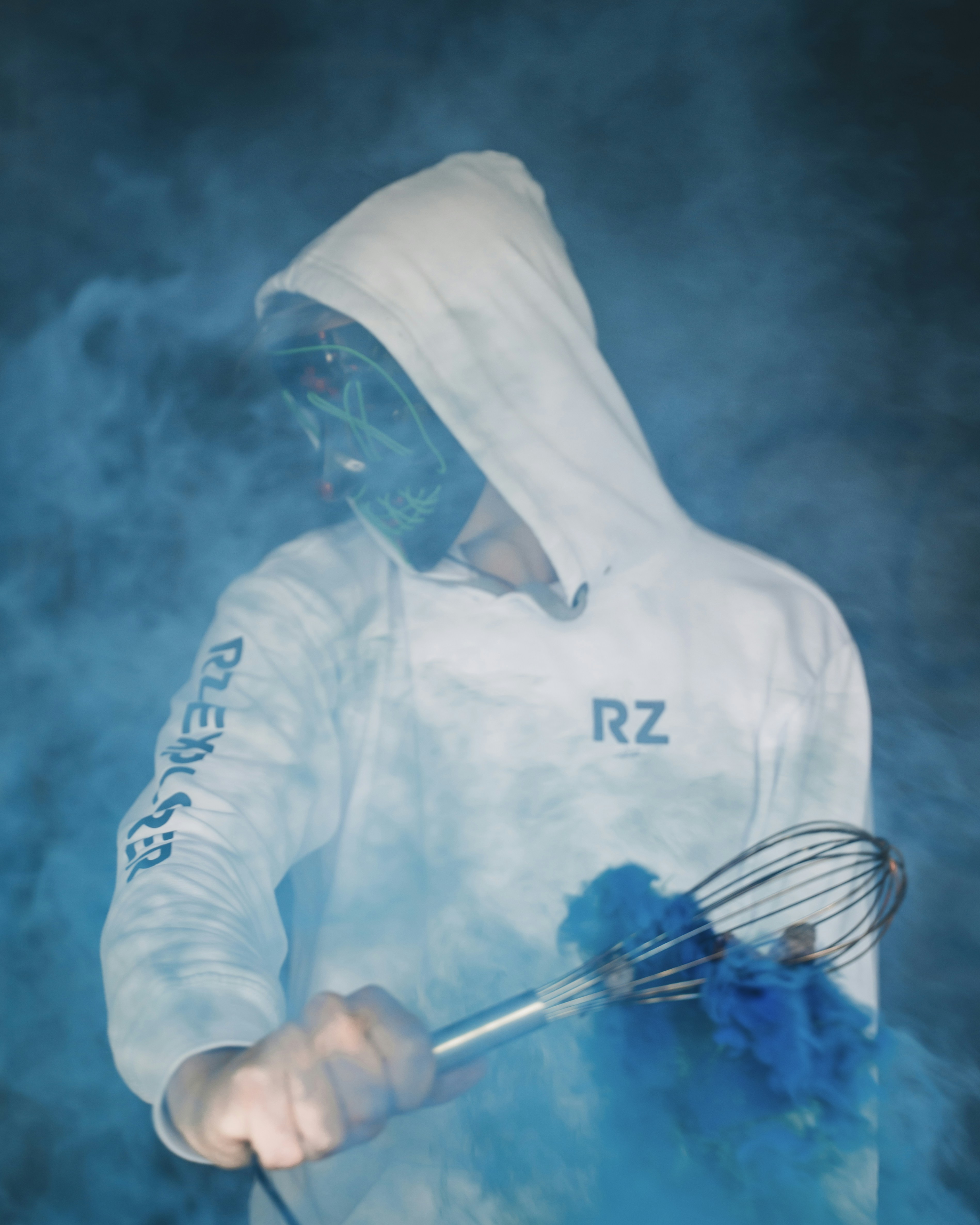 A figure in a white hoodie and face paint wields a whisk amidst swirling blue smoke, creating an intriguing atmosphere of creativity and mystery.