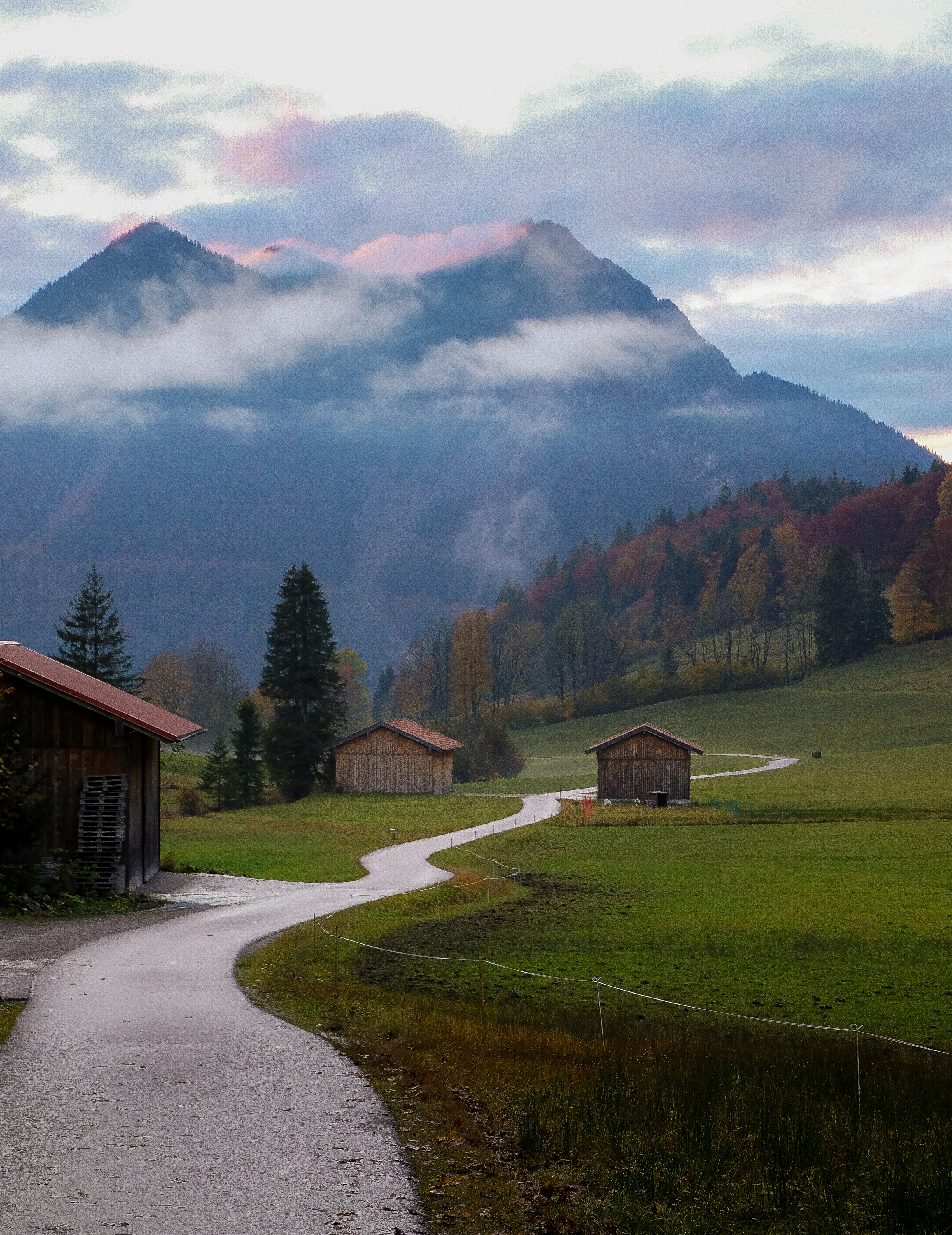 Curved road leads through a serene landscape dotted with rustic wooden cabins and vibrant autumn foliage against a backdrop of majestic mountains.