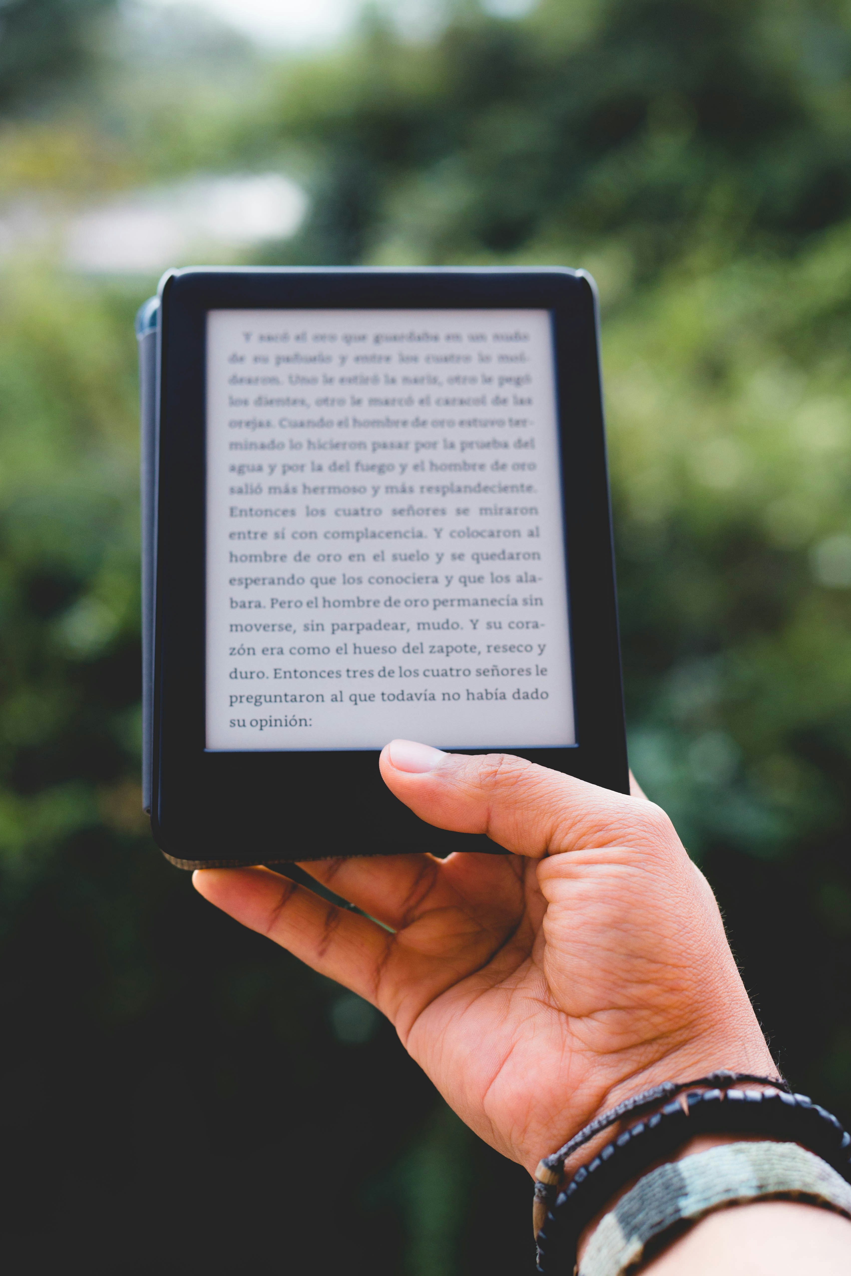 A hand holds a black e-reader displaying a block of text in Spanish. The device is photographed against a soft, leafy green background.