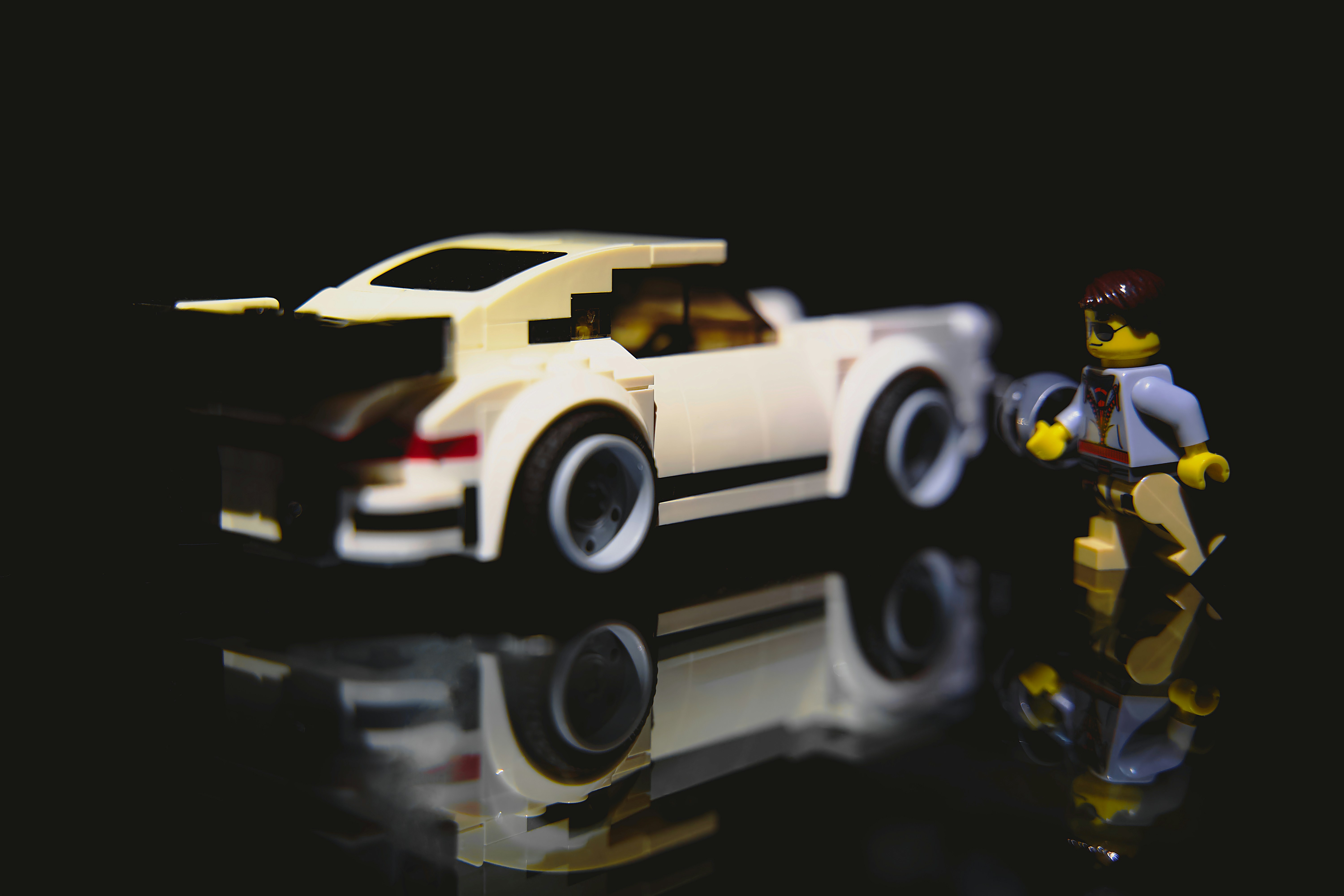 A LEGO figure stealthily approaches a white sports car, set against a reflective black background, creating a playful scene of intrigue.