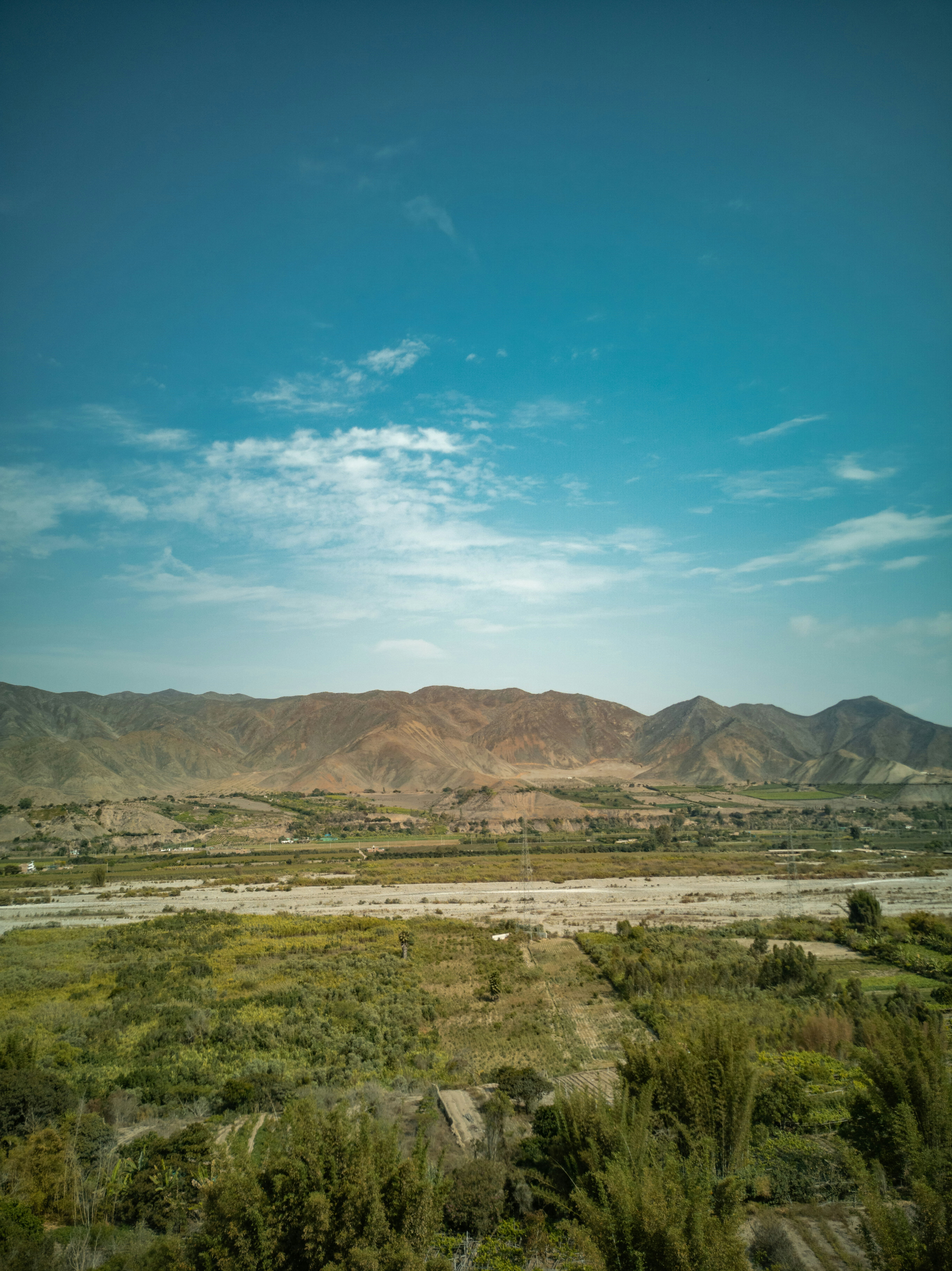 Landscape photograph of a valley with scrubby vegetation and a distant mountain ridge under a bright blue sky.
