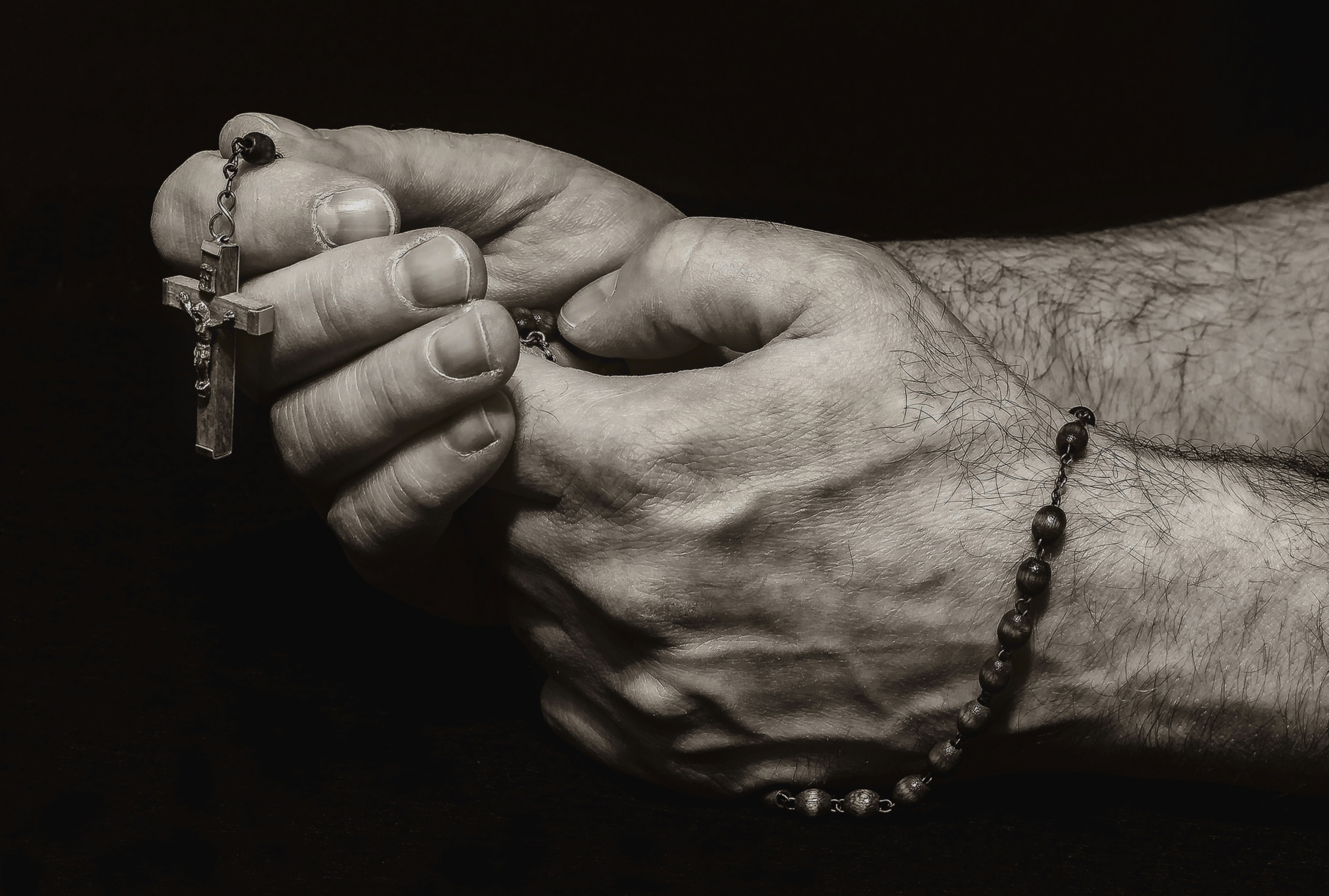 praying hands on black background | grayscale photo of person holding silver necklace