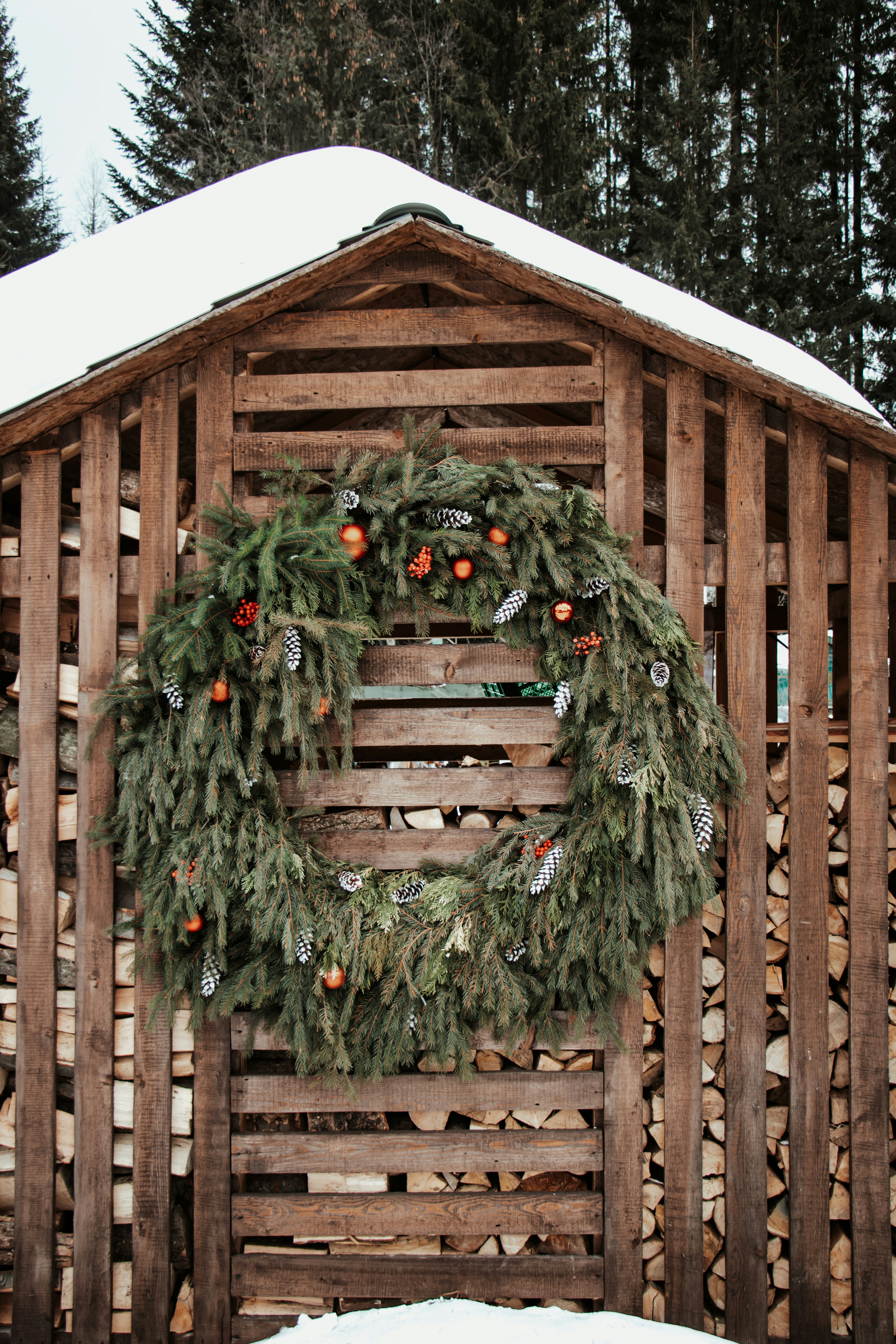 A beautifully adorned evergreen wreath featuring pinecones and berries, mounted on a rustic wooden backdrop. The scene evokes a warm, festive atmosphere.