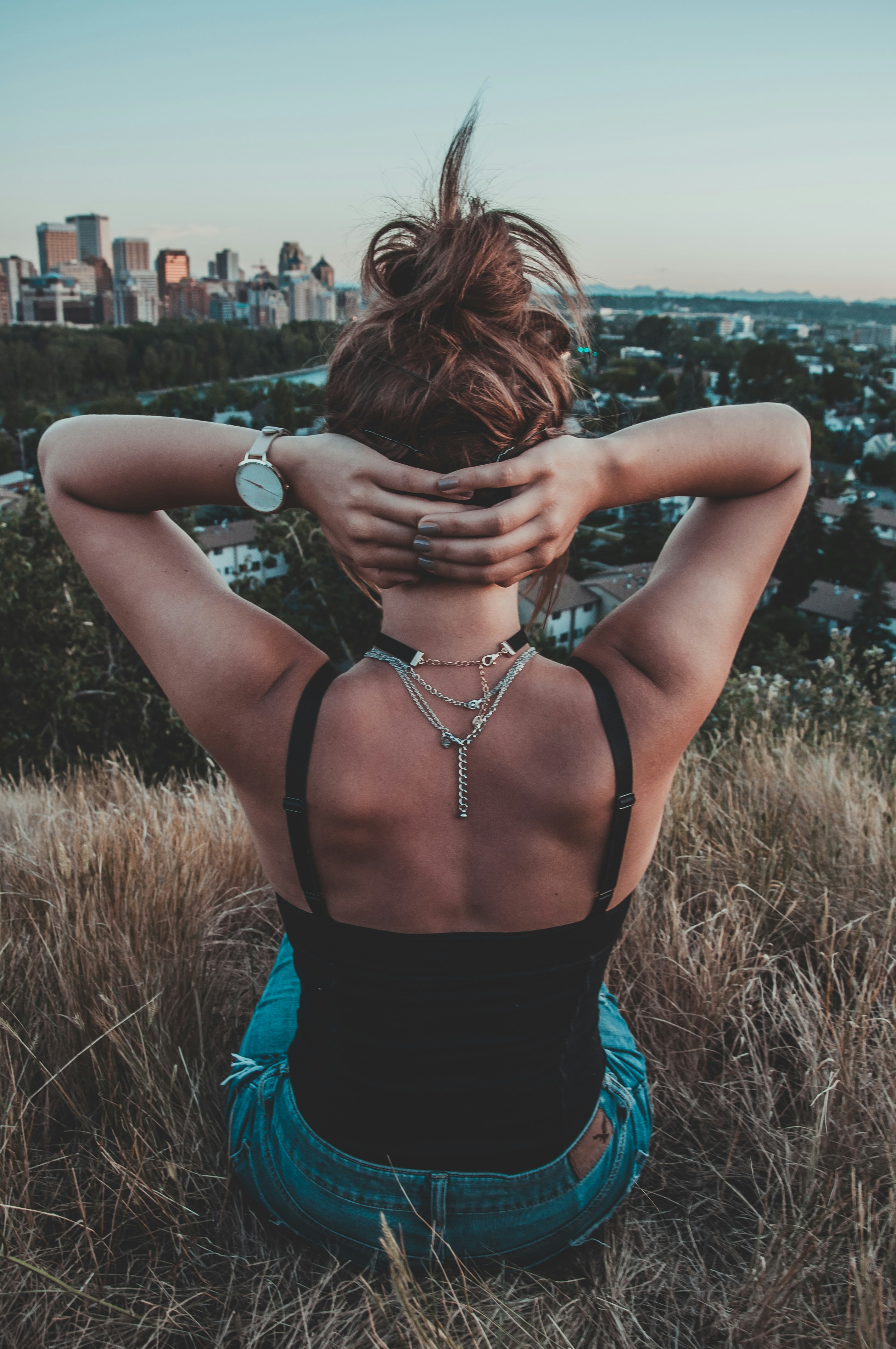 A woman sitting in a grassy area, gazing at a city skyline during twilight, with her hair styled up and adorned with layered jewelry.