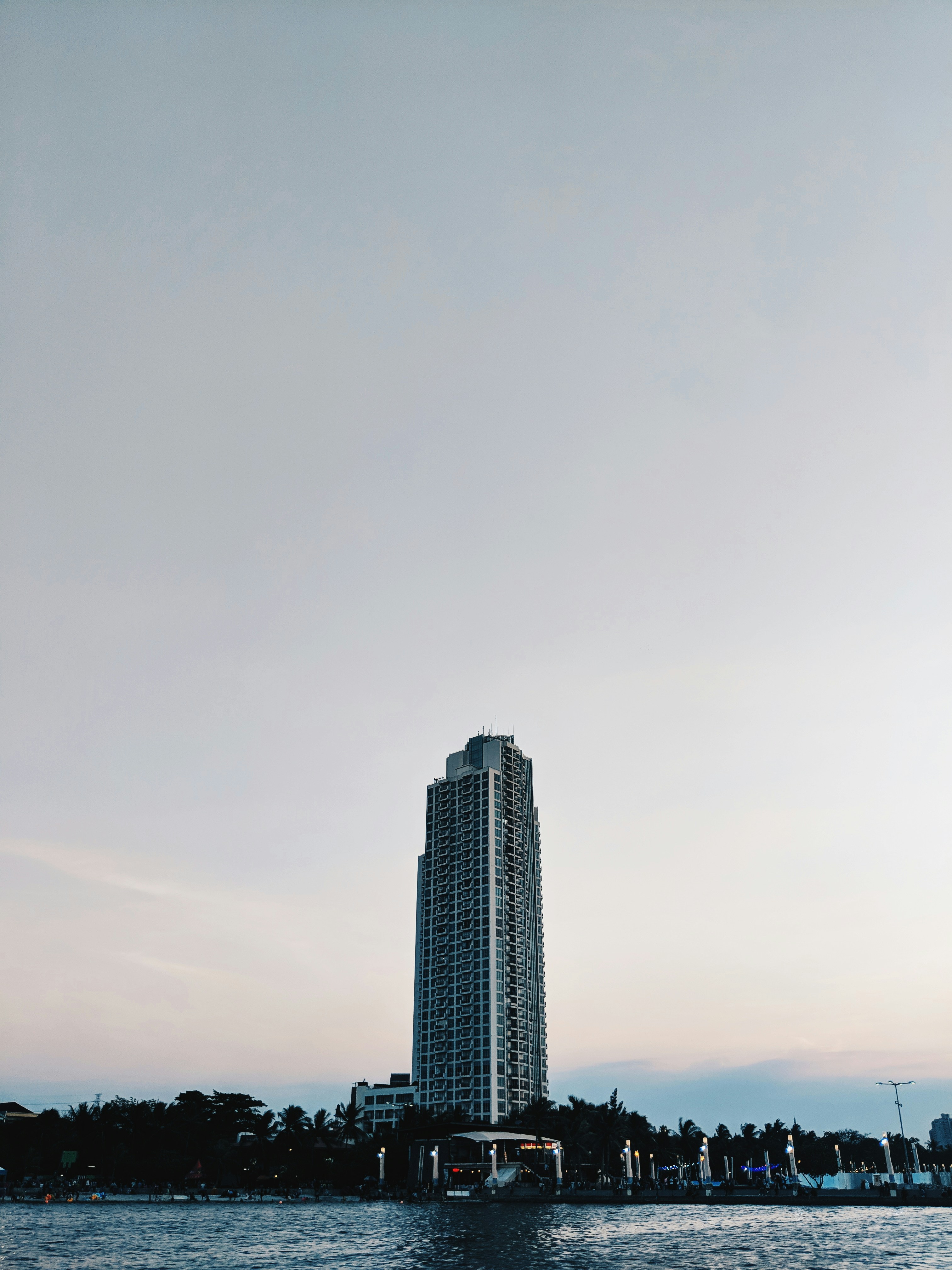 A tall modern skyscraper stands against a pastel sky, reflecting in the water below, surrounded by lush greenery and urban life.
